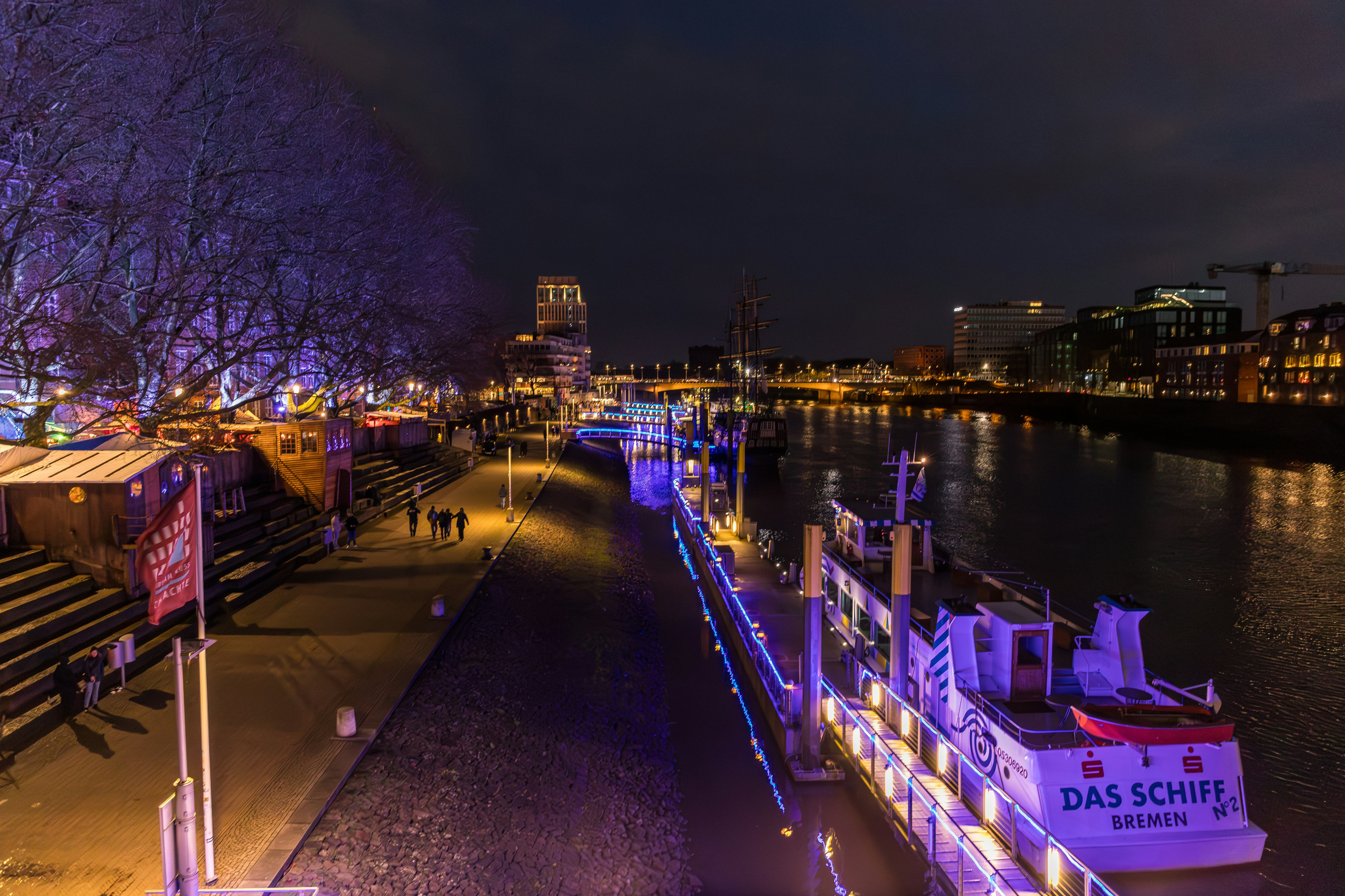 Vibrant lights illuminating a riverside promenade at night, with boats docked along the water's edge. The scene captures the lively atmosphere of an urban waterfront.
