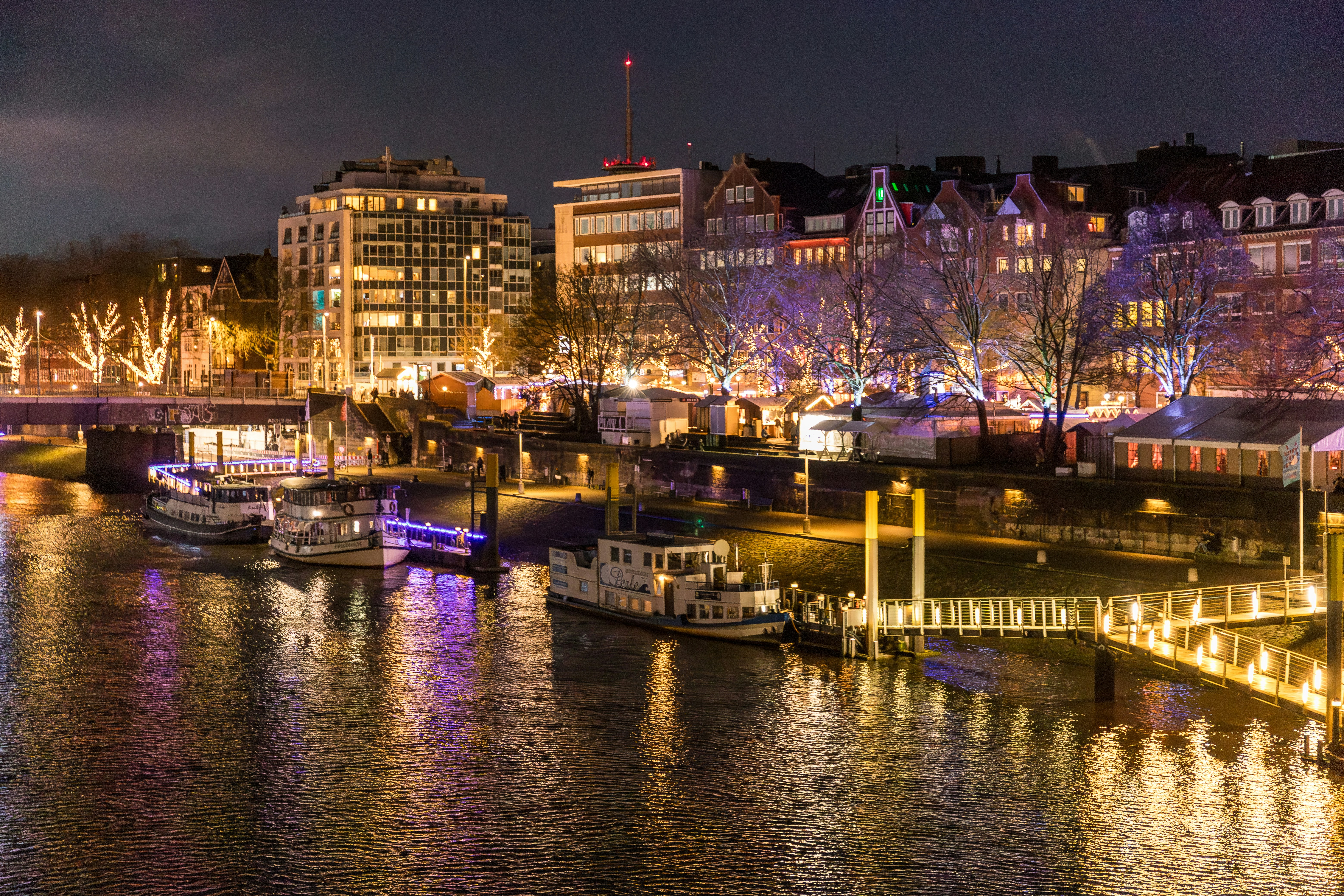 City harbor at night with boats docked under vibrant city lights.