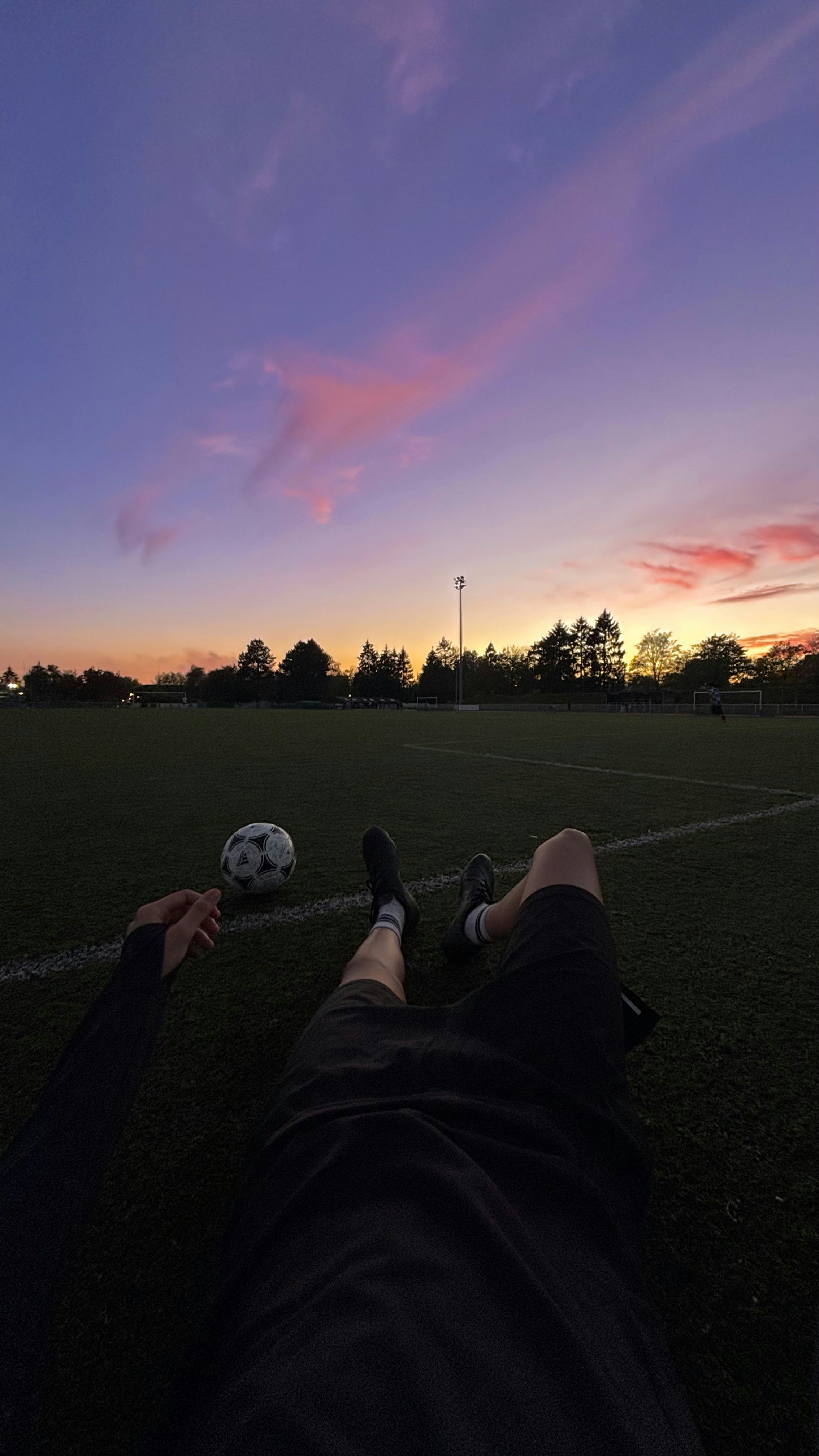 A player reclines on a soccer field at dusk, with a ball nearby and a vibrant sunset illuminating the sky. The scene captures the tranquility of post-game moments.