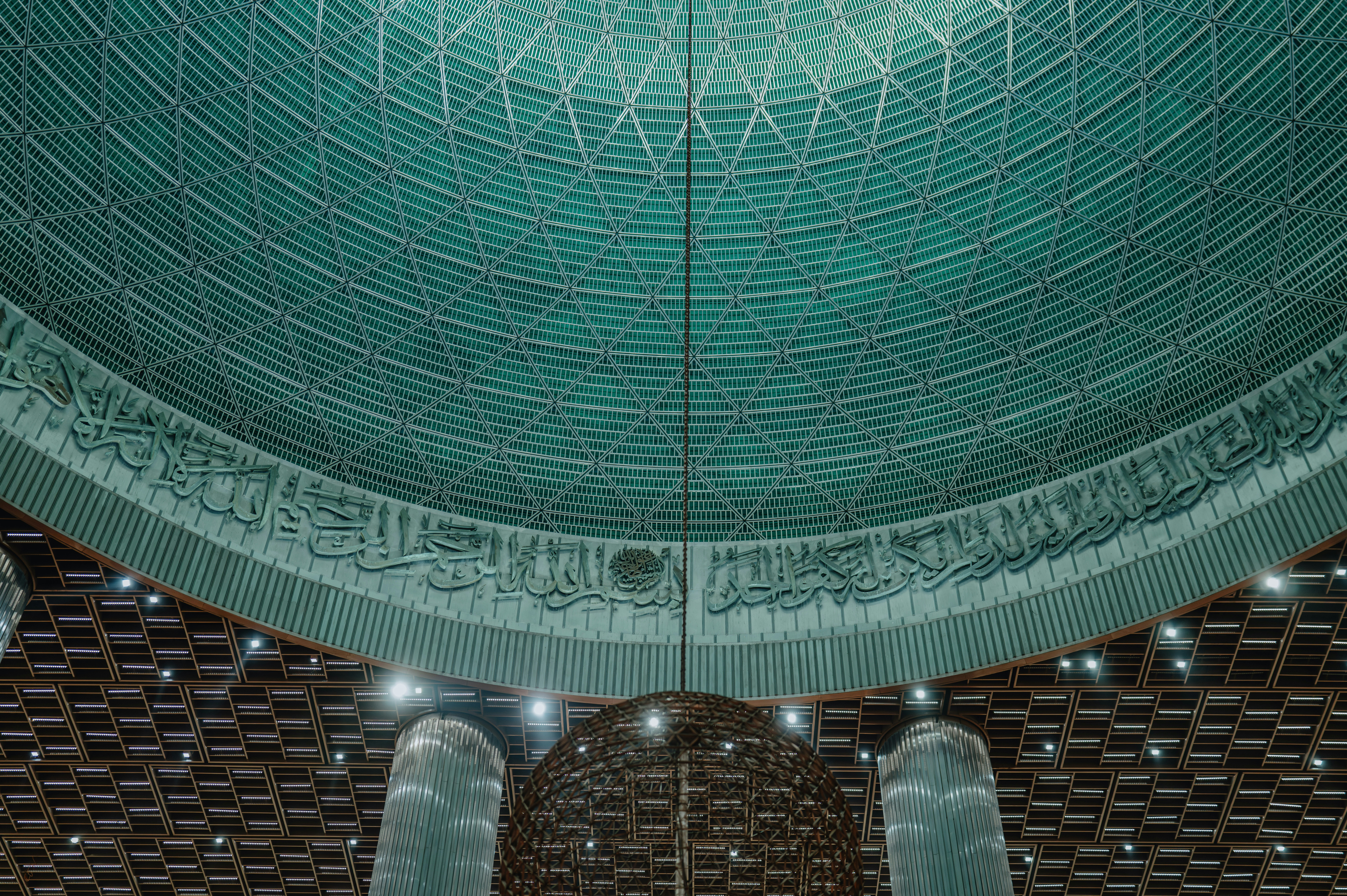 Teal-dominated architectural interior photograph featuring a symmetrical domed ceiling with an intricate lattice grid, flanked by massive columns and decorative arches.