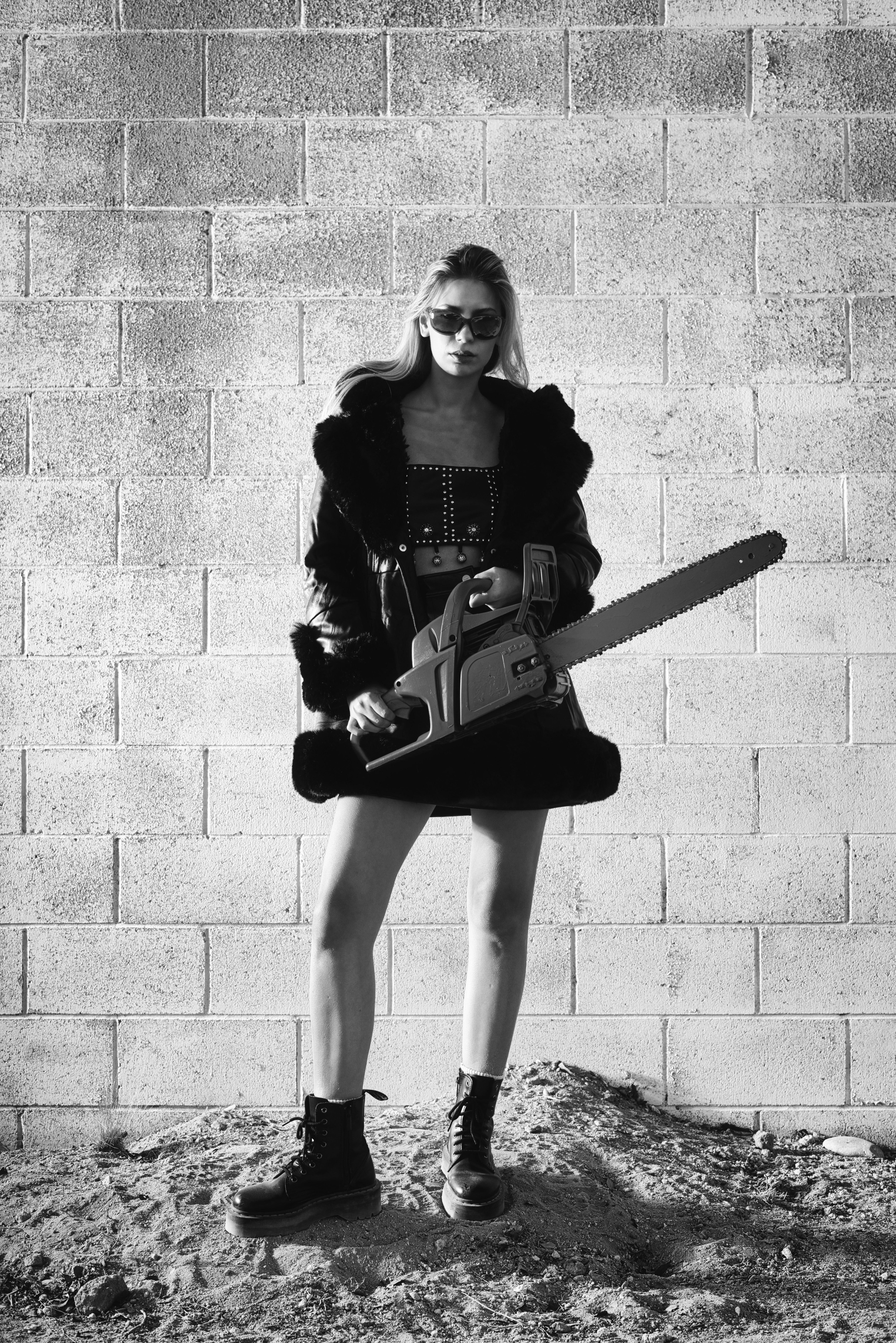 Model in a stylish fur coat and sunglasses holds a chainsaw against a textured concrete wall, conveying a striking contrast of fashion and industrial elements.