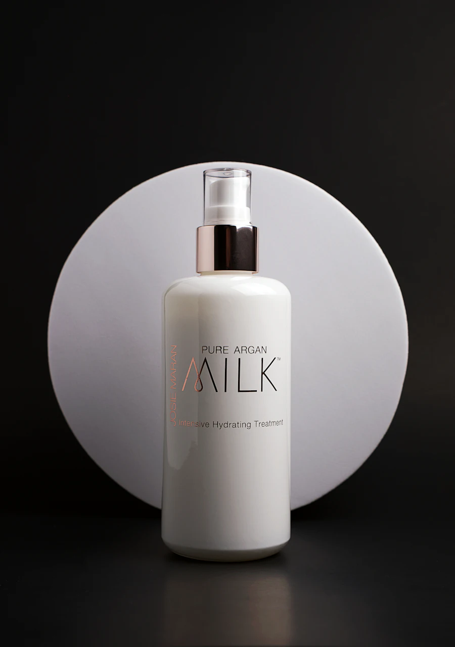 White Pure Argan Milk hair treatment bottle centered in front of a pale circular backdrop on a black background. Soft studio lighting highlights the product and its reflective pump top.