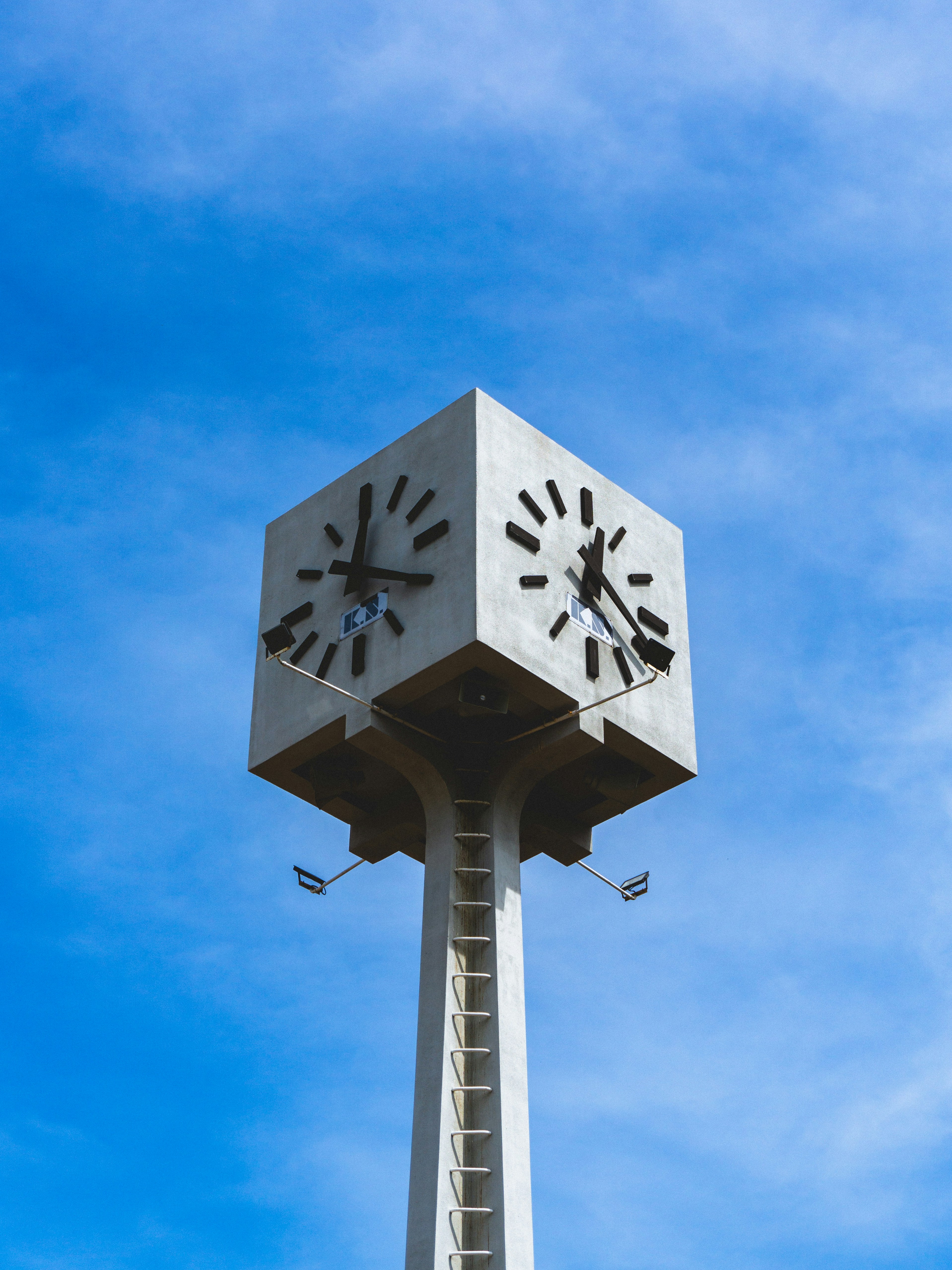 A modern concrete clock tower with distinct clock faces stands tall against a clear blue sky.