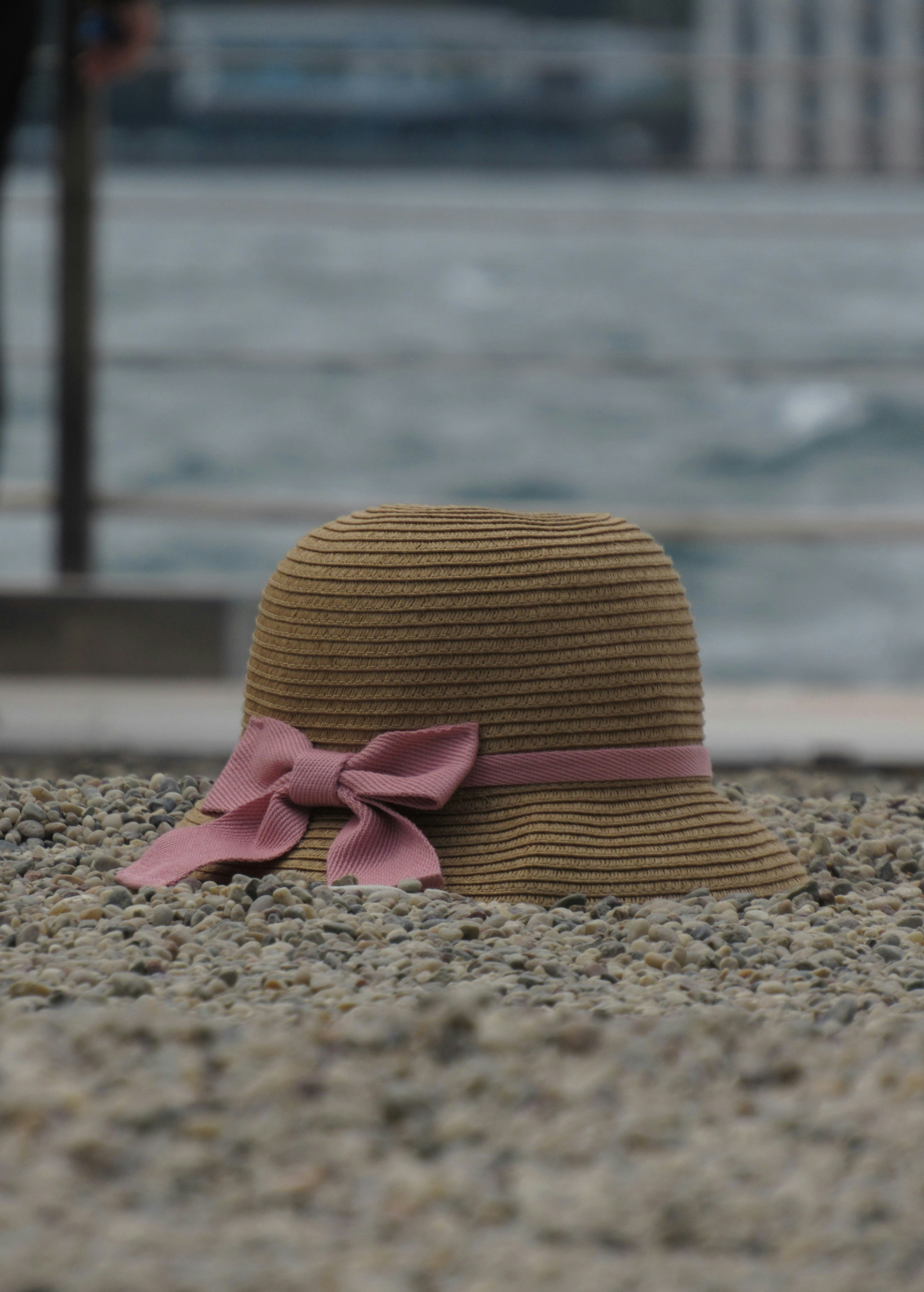 Straw hat with a pink ribbon rests on a pebbled shore, with blurred water and railing in the background. The still life emphasizes texture and contrast between the hat and the gritty foreground.