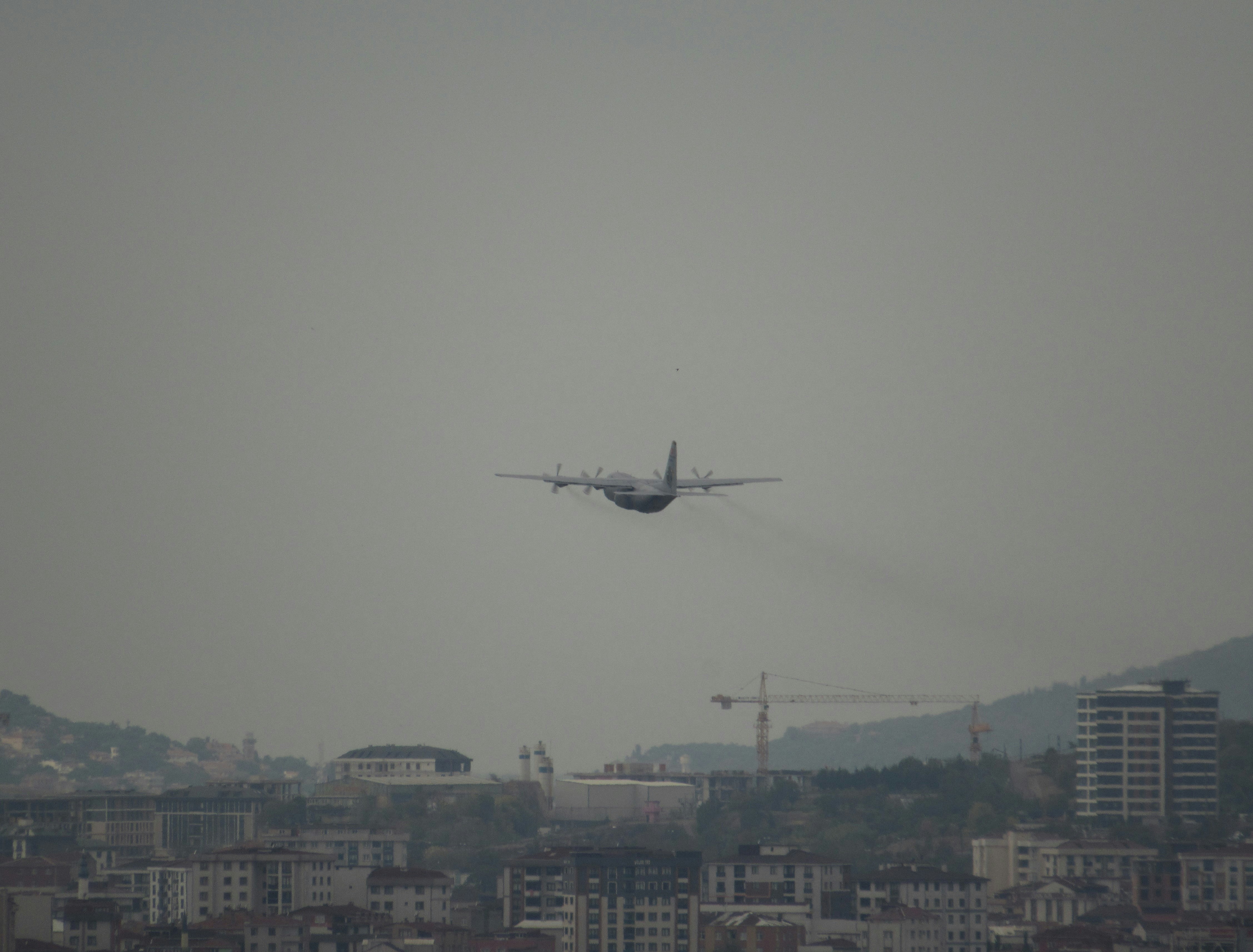 A military transport plane, likely a C-130 Hercules, is captured in mid-flight, ascending above a cityscape. The plane's distinctive silhouette and powerful engines are clearly visible against the overcast sky. The cityscape in the background provides a sense of scale and context, emphasizing the aircraft's size and capabilities.