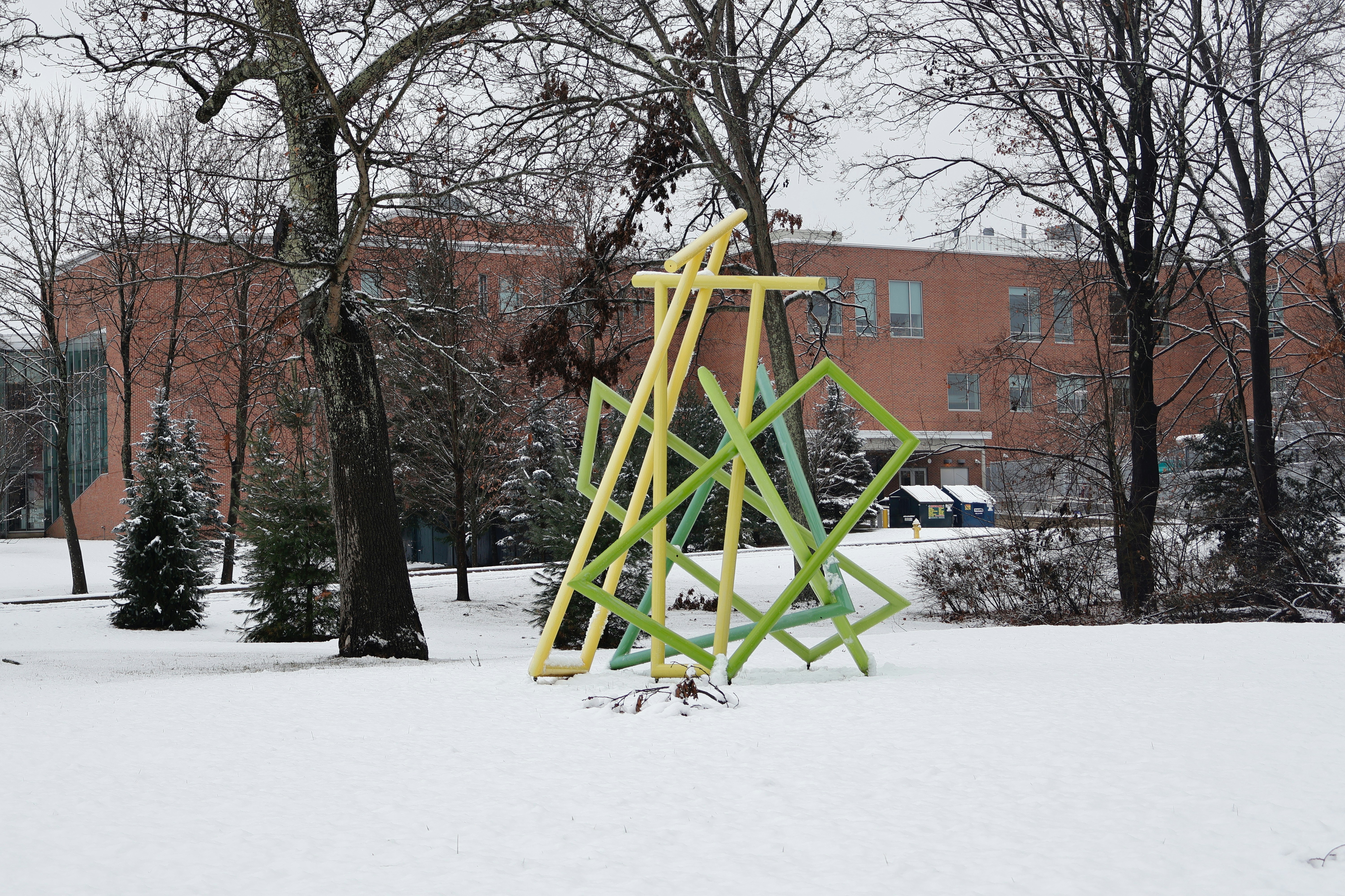 Colorful abstract sculpture made of green and yellow geometric shapes standing in a snowy landscape with trees in the background.
