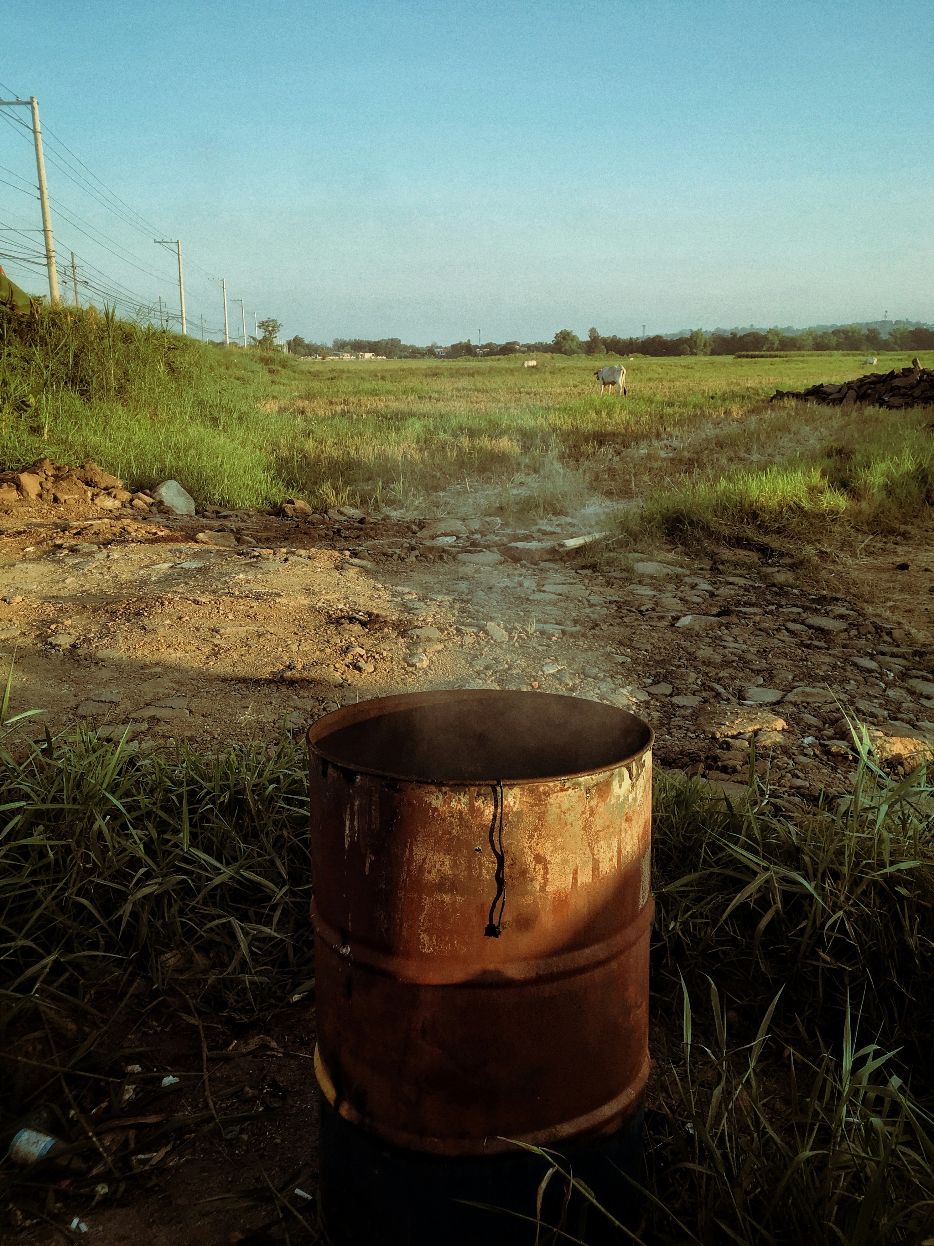 Rust-bucket at the Edge: Smoke Rises from Iron Barrel in a Dusty Field