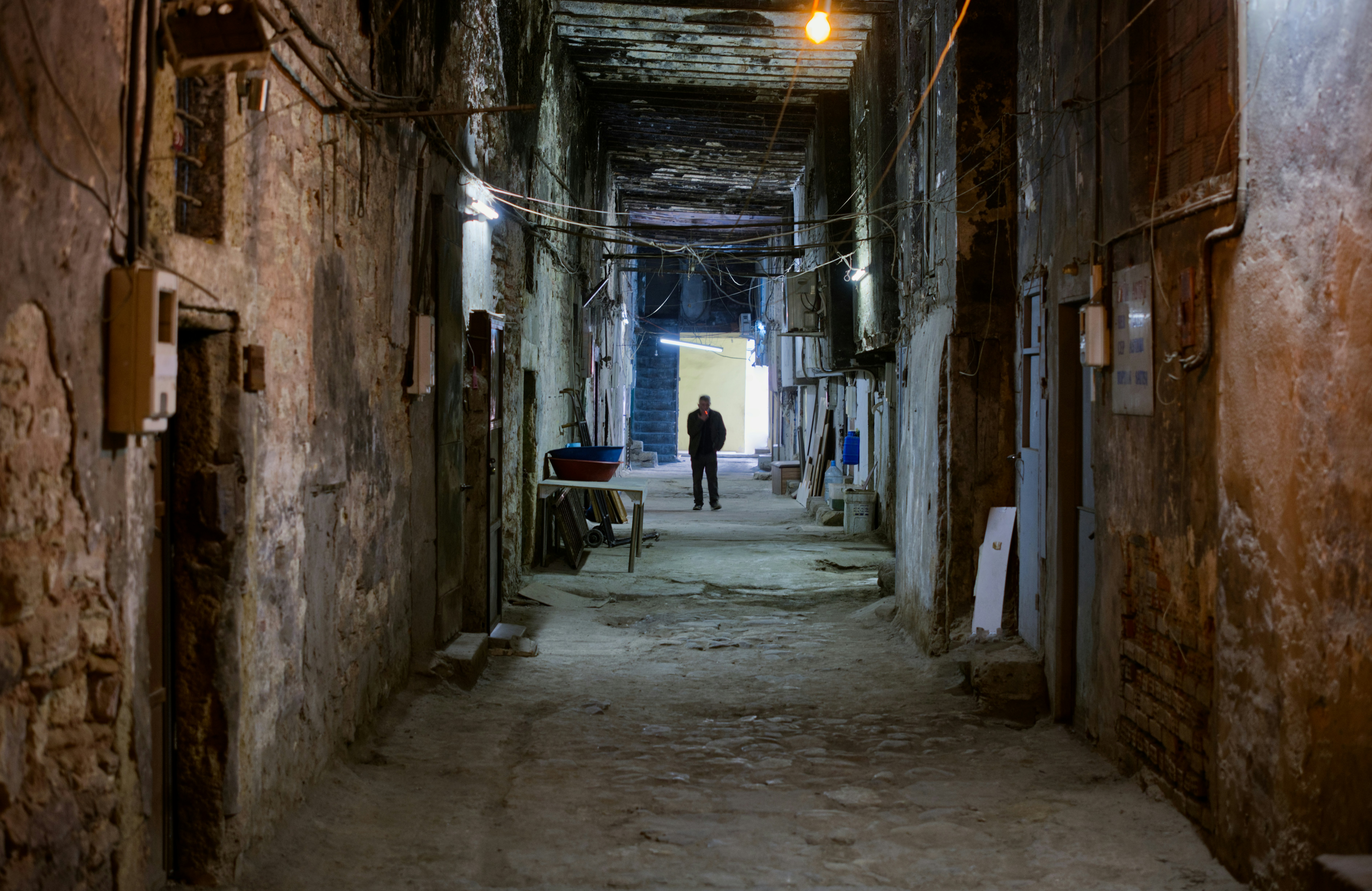 Dimly lit corridor with weathered walls and a solitary figure walking towards a bright exit.