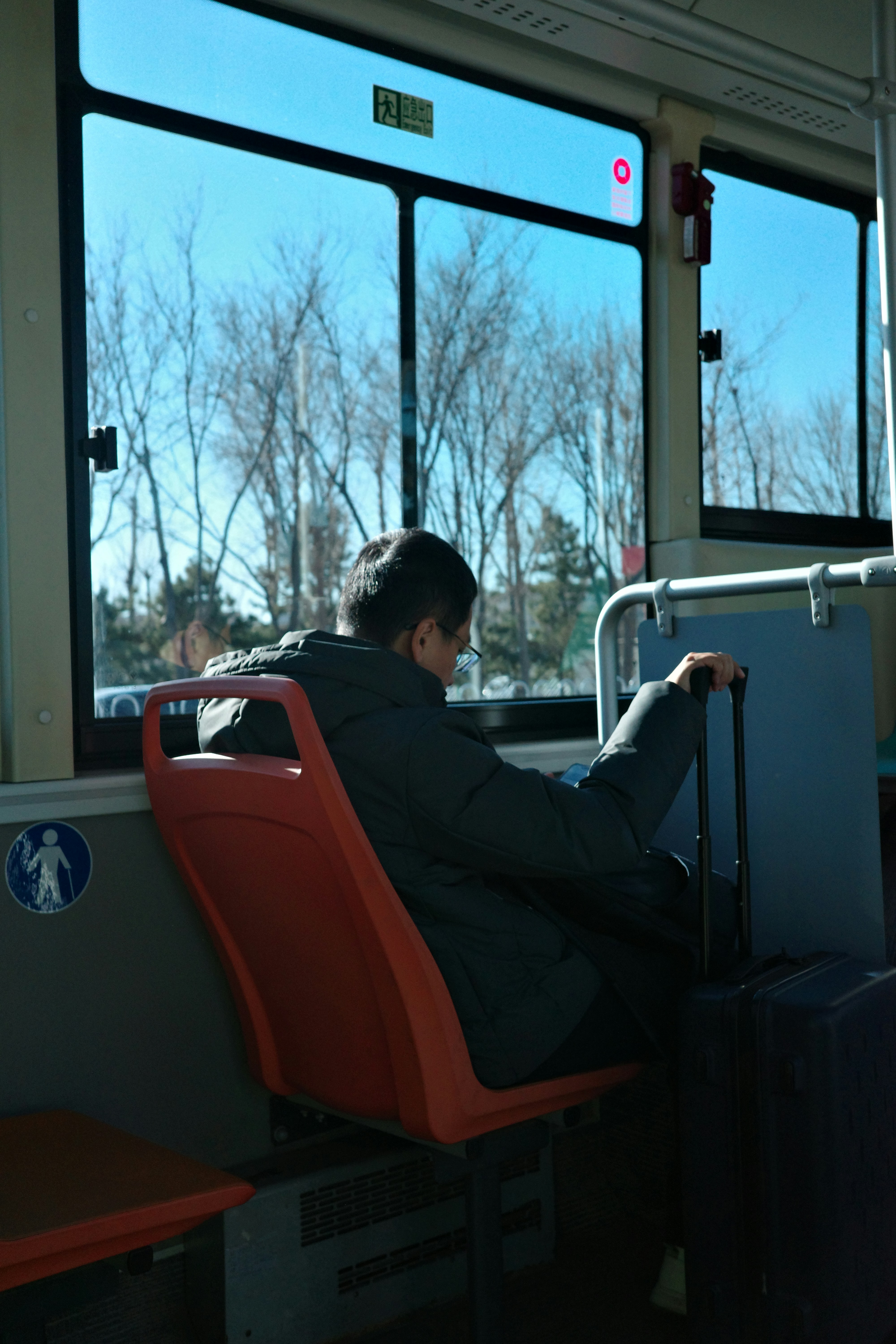A passenger seated on a bus, lost in thought, with a suitcase beside them and sunlight streaming through the window.