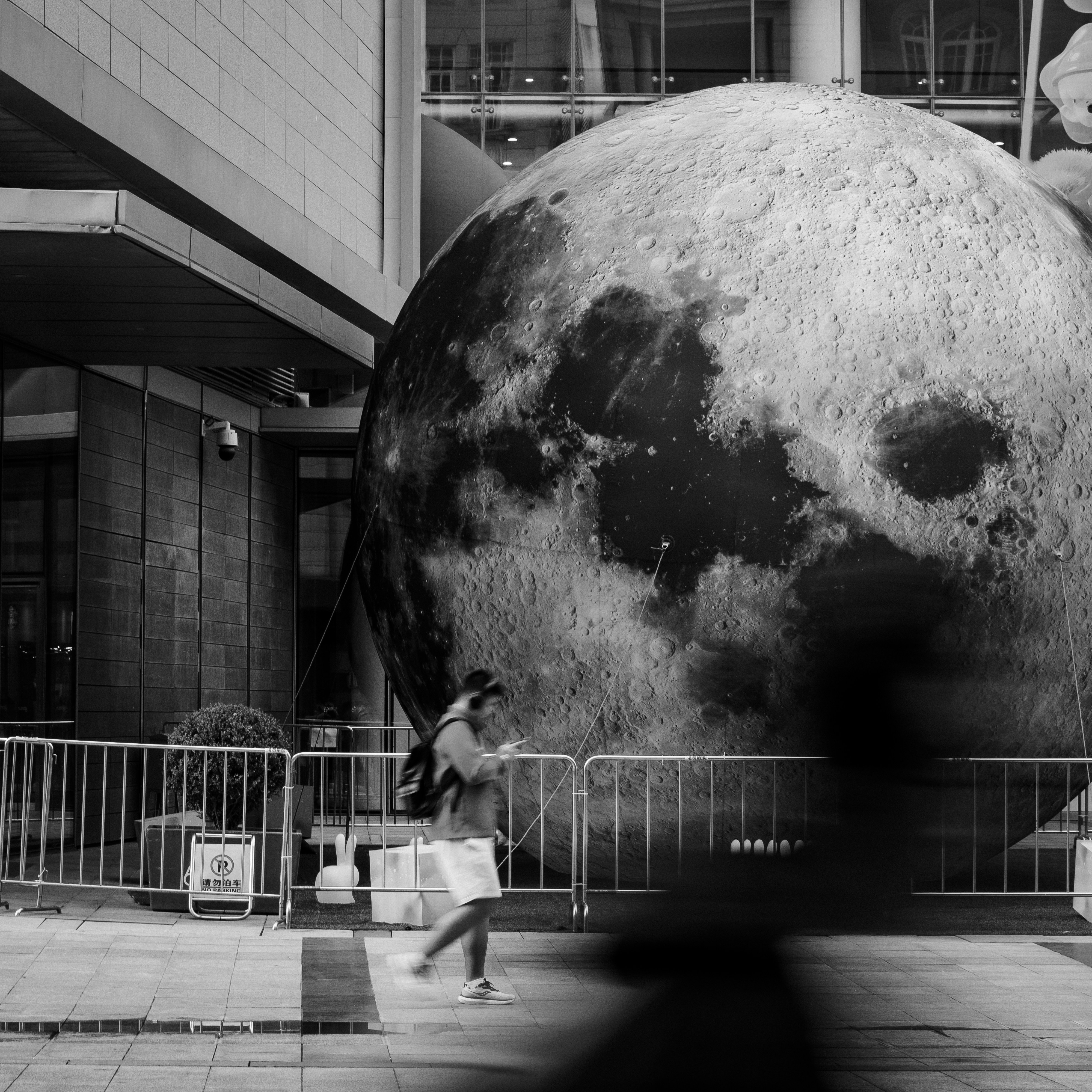 Moon over Motion: A Pedestrian Passes Before a Giant Lunar Sculpture