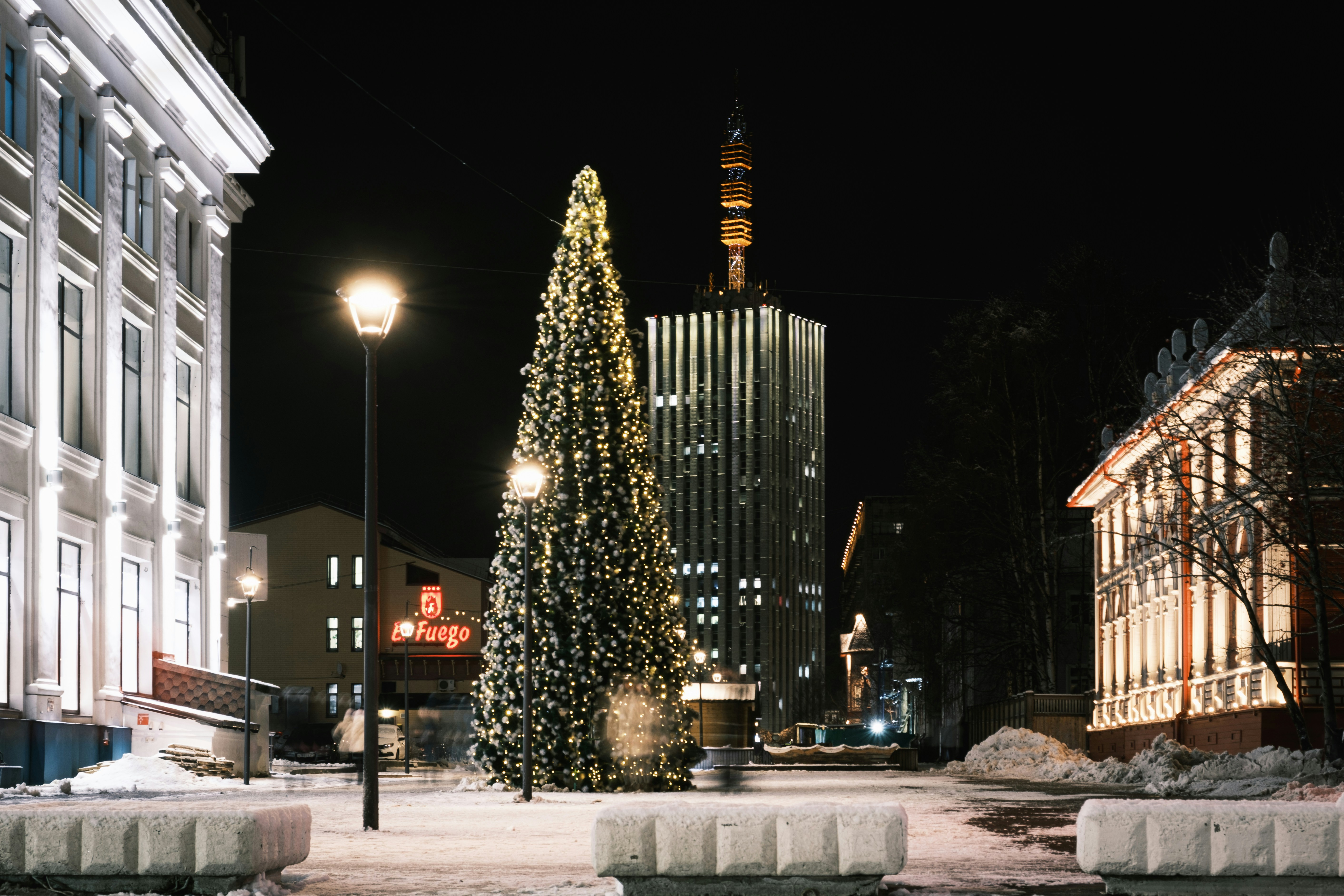 Illuminated Christmas tree set amidst city architecture under night sky.