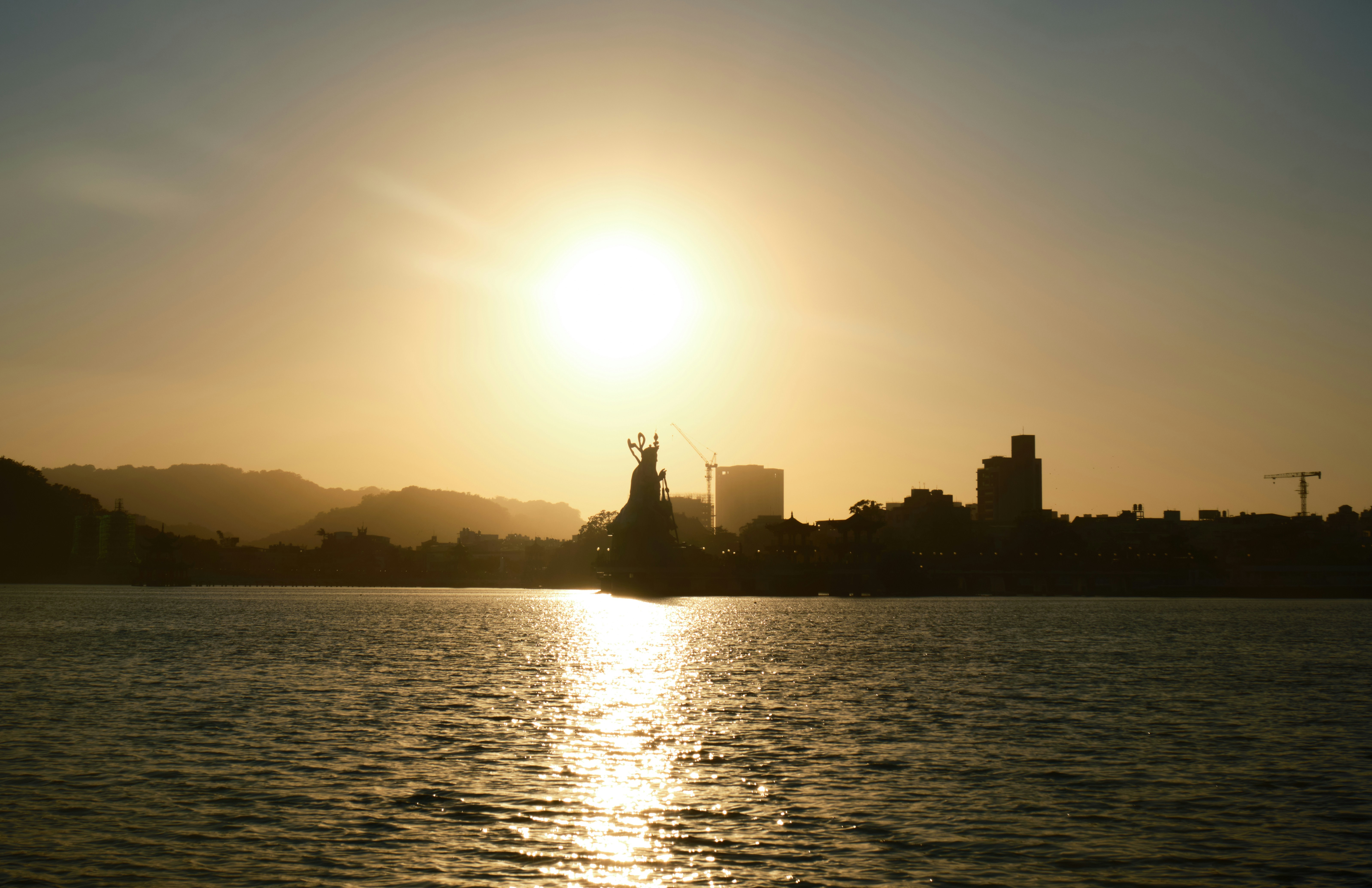 Golden sun setting behind a city's skyline, casting shimmering reflections on a calm body of water.