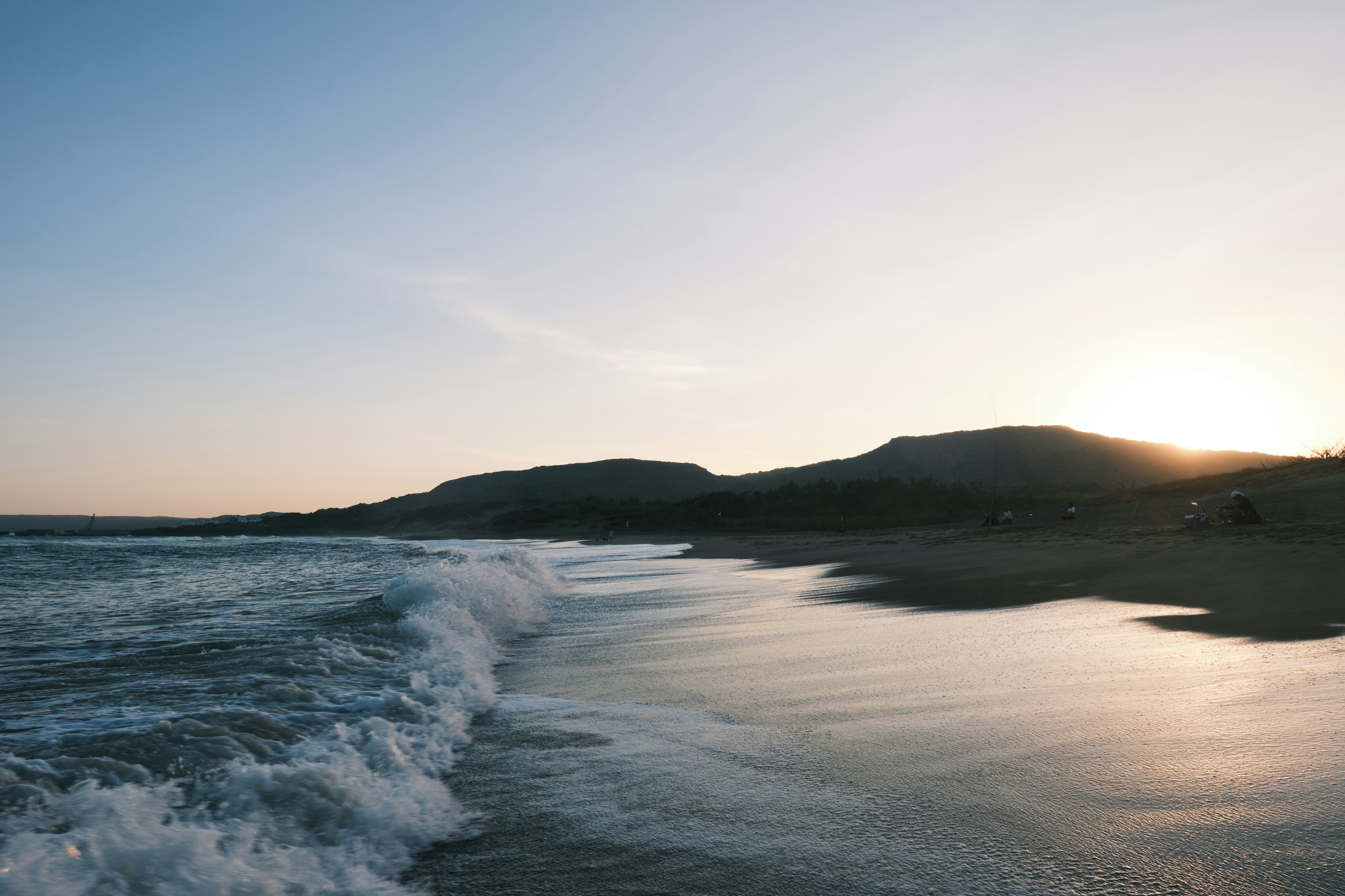Gentle waves roll onto a sandy beach as the sun sets behind distant hills.