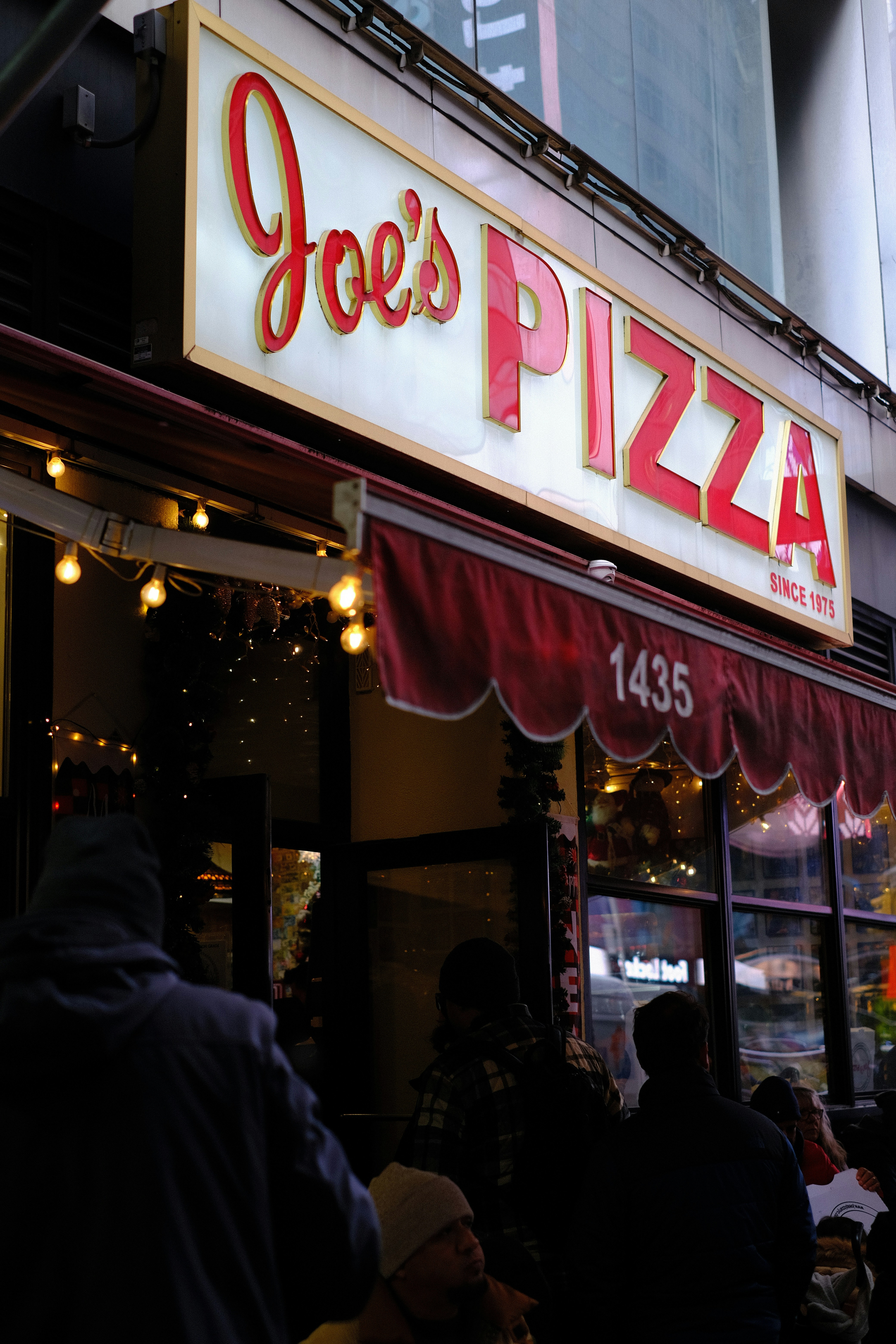 Neon sign for Joe's Pizza with a red awning and festive string lights, capturing a lively urban street scene.