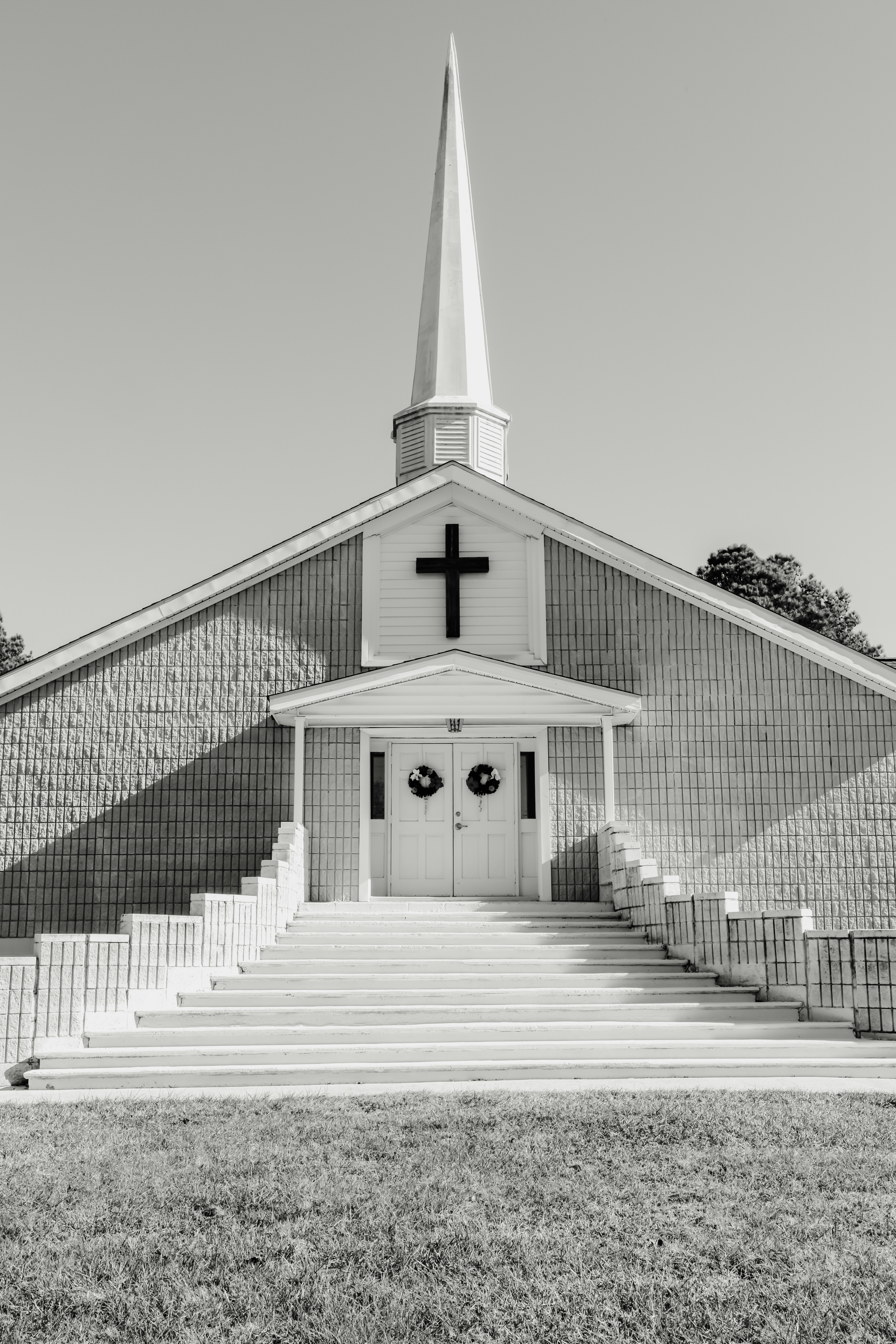 Black and white photograph of a church facade with a tall steeple and cross, featuring strong architectural lines and shadows on textured brickwork.