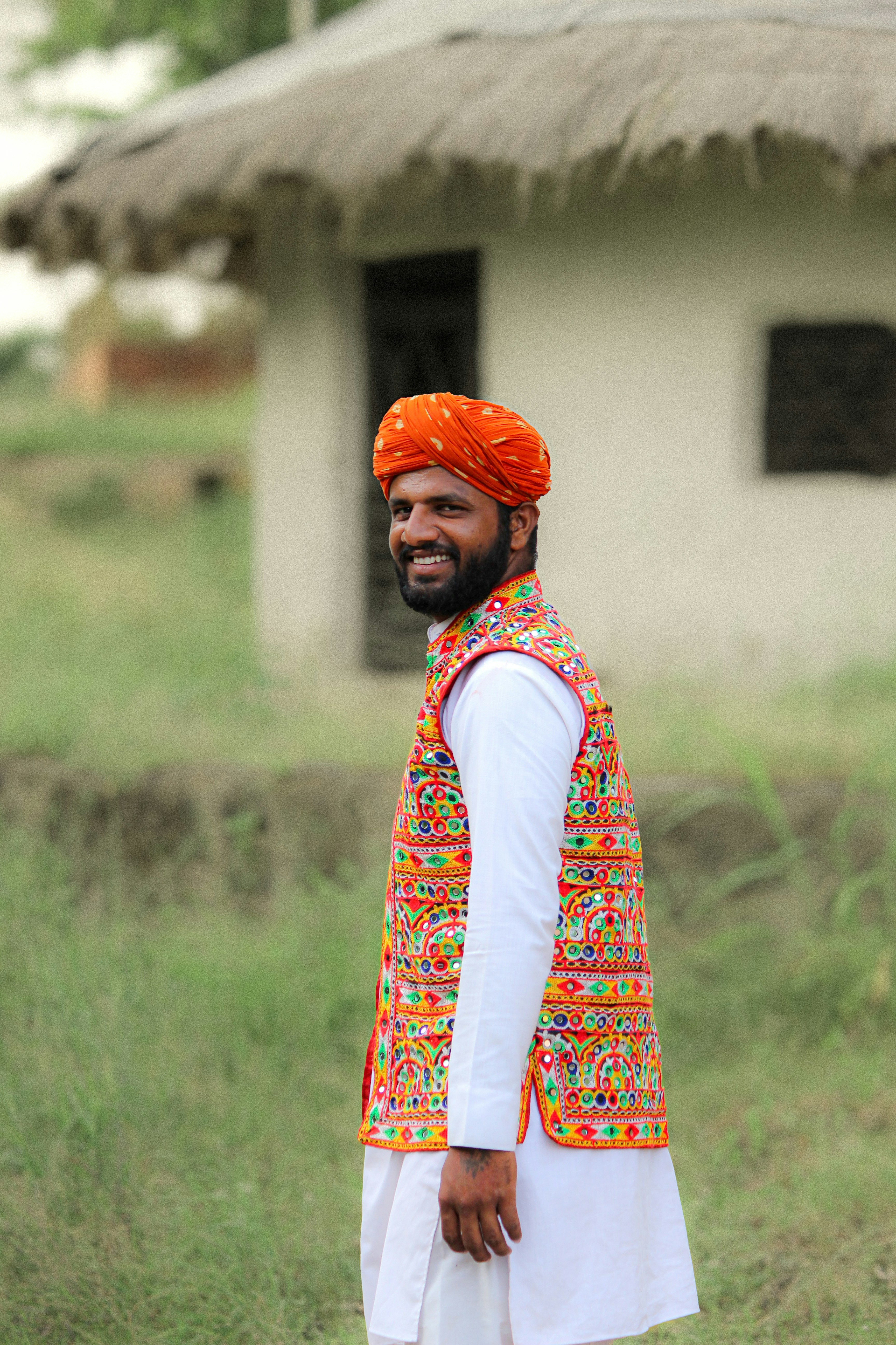 Man in a colorful traditional outfit and turban standing near a thatched hut in a grassy field.
