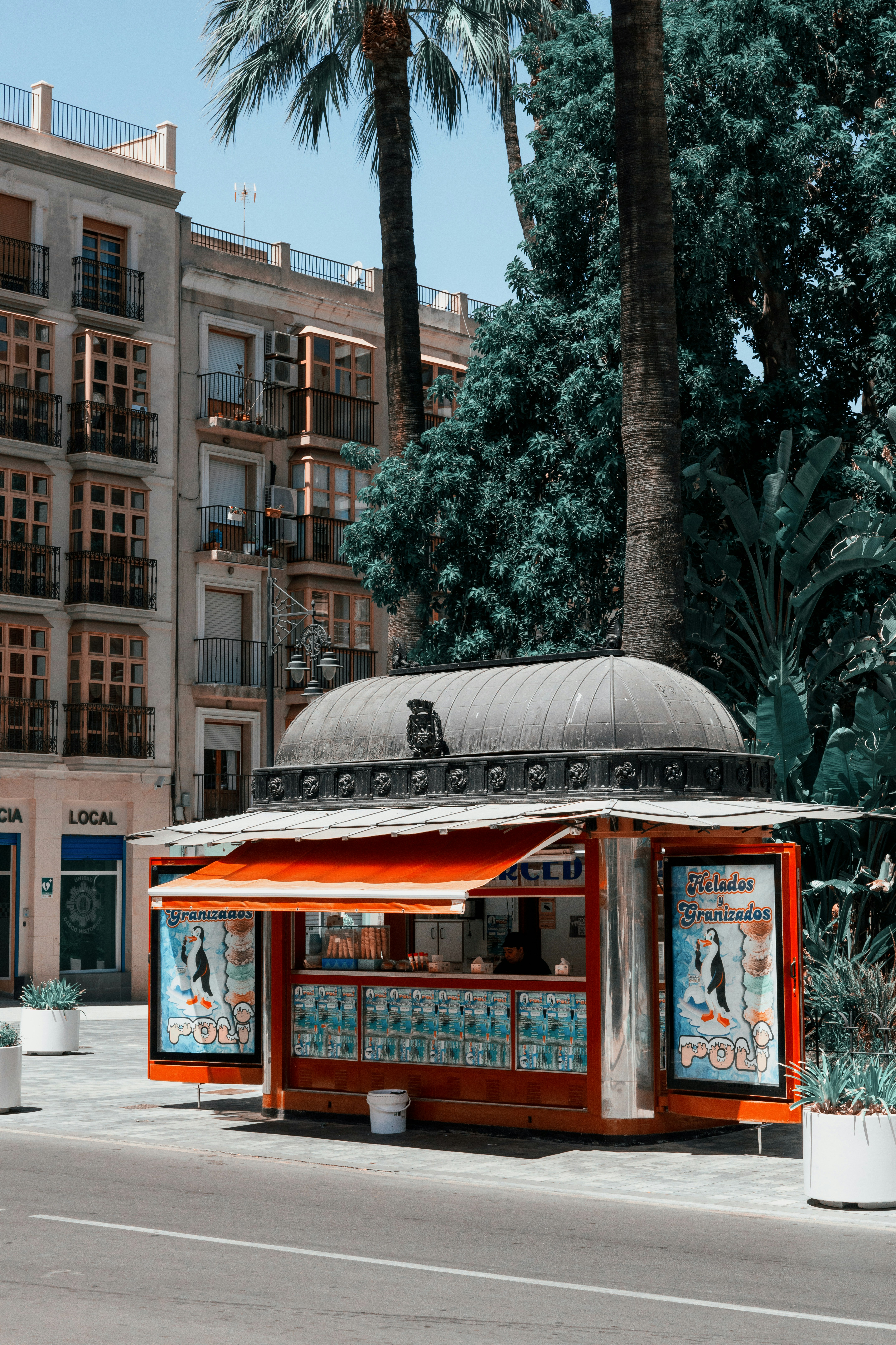Orange kiosk under a domed canopy amid palm trees and city balconies