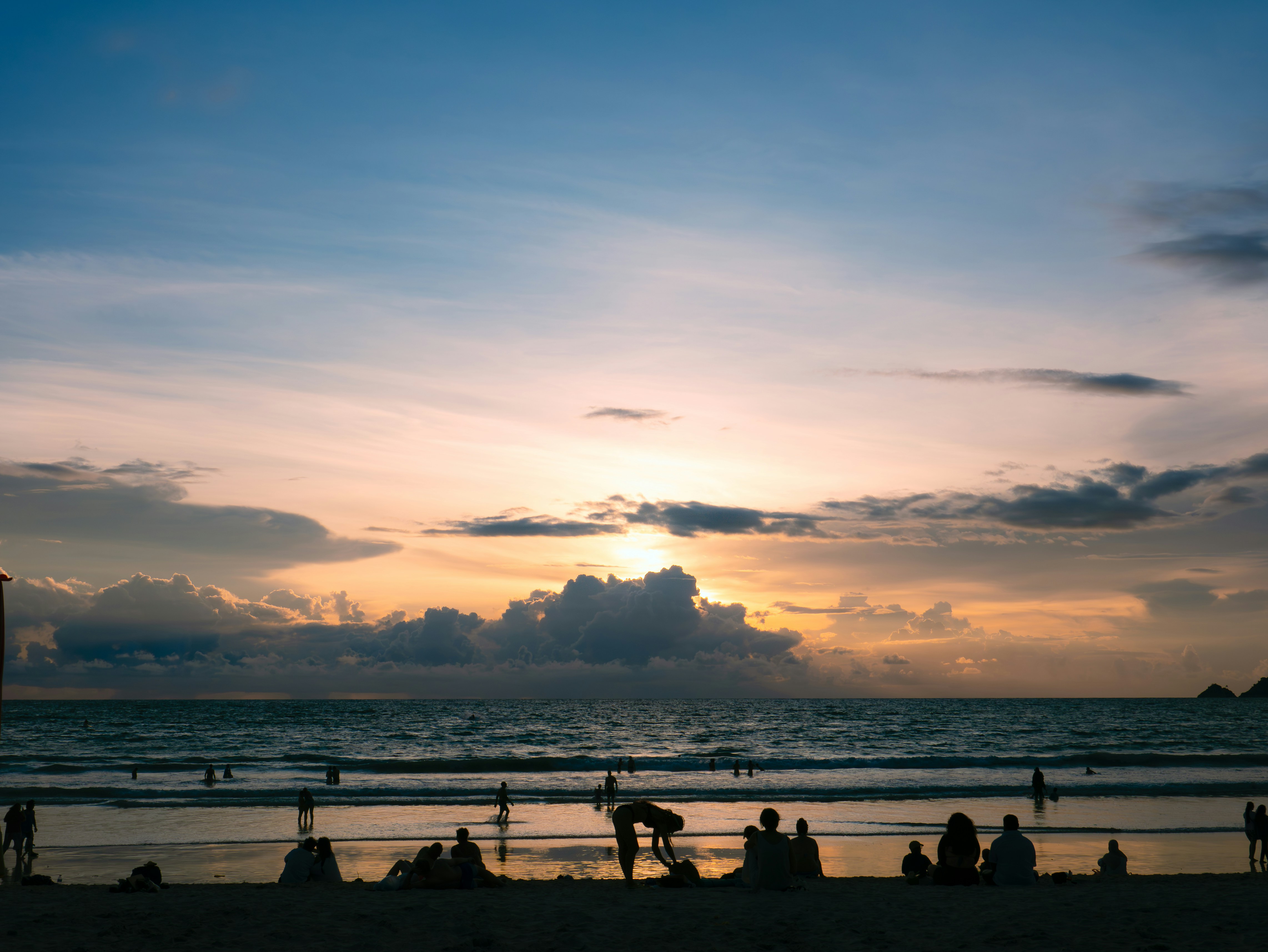 Silhouetted figures against a vibrant sunset over the ocean, with clouds scattered across the sky.