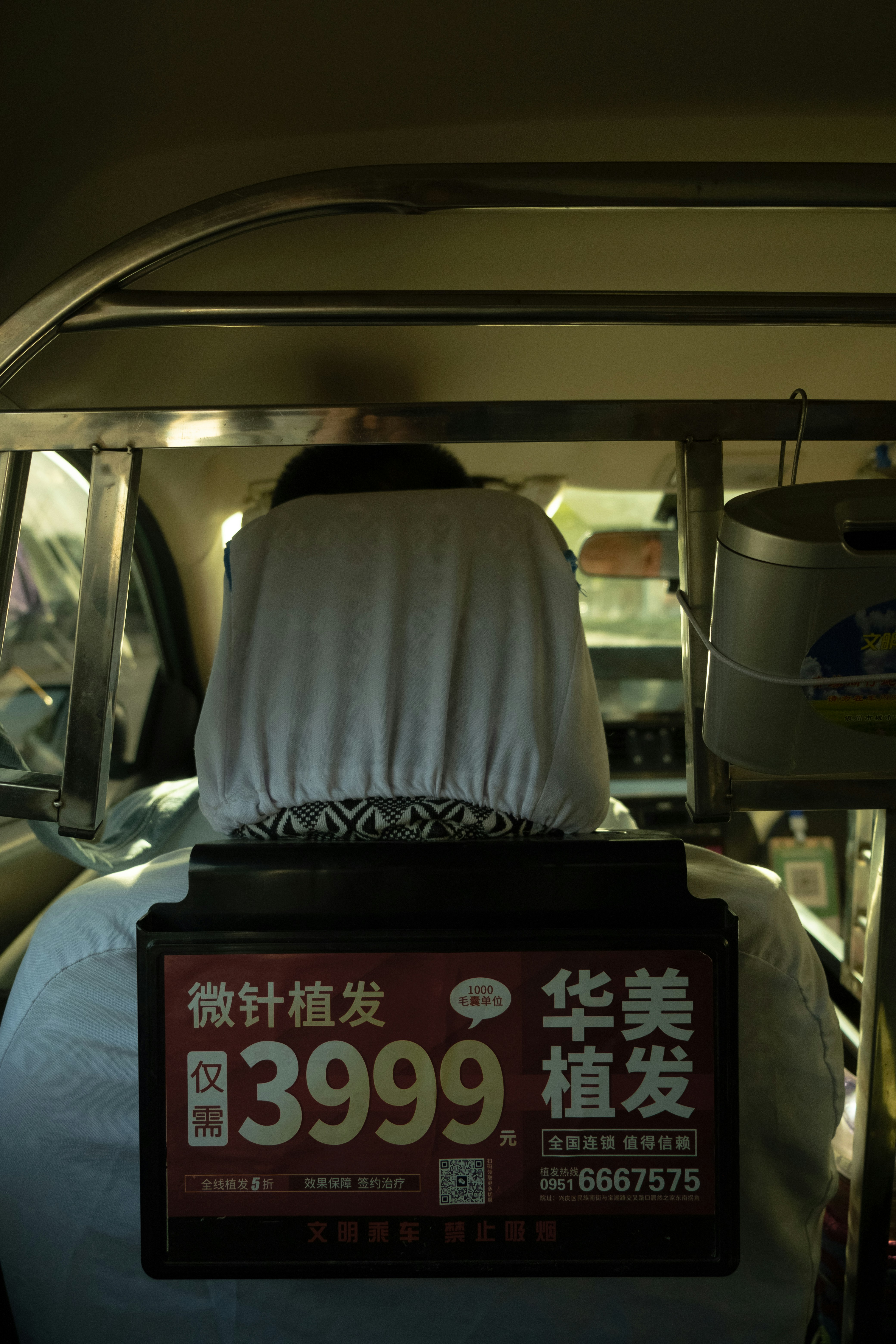 Dim taxi interior features a back-of-seat red advertisement board displaying a large '3999' price for hair extensions, with a white cloth covering the driver's seat.