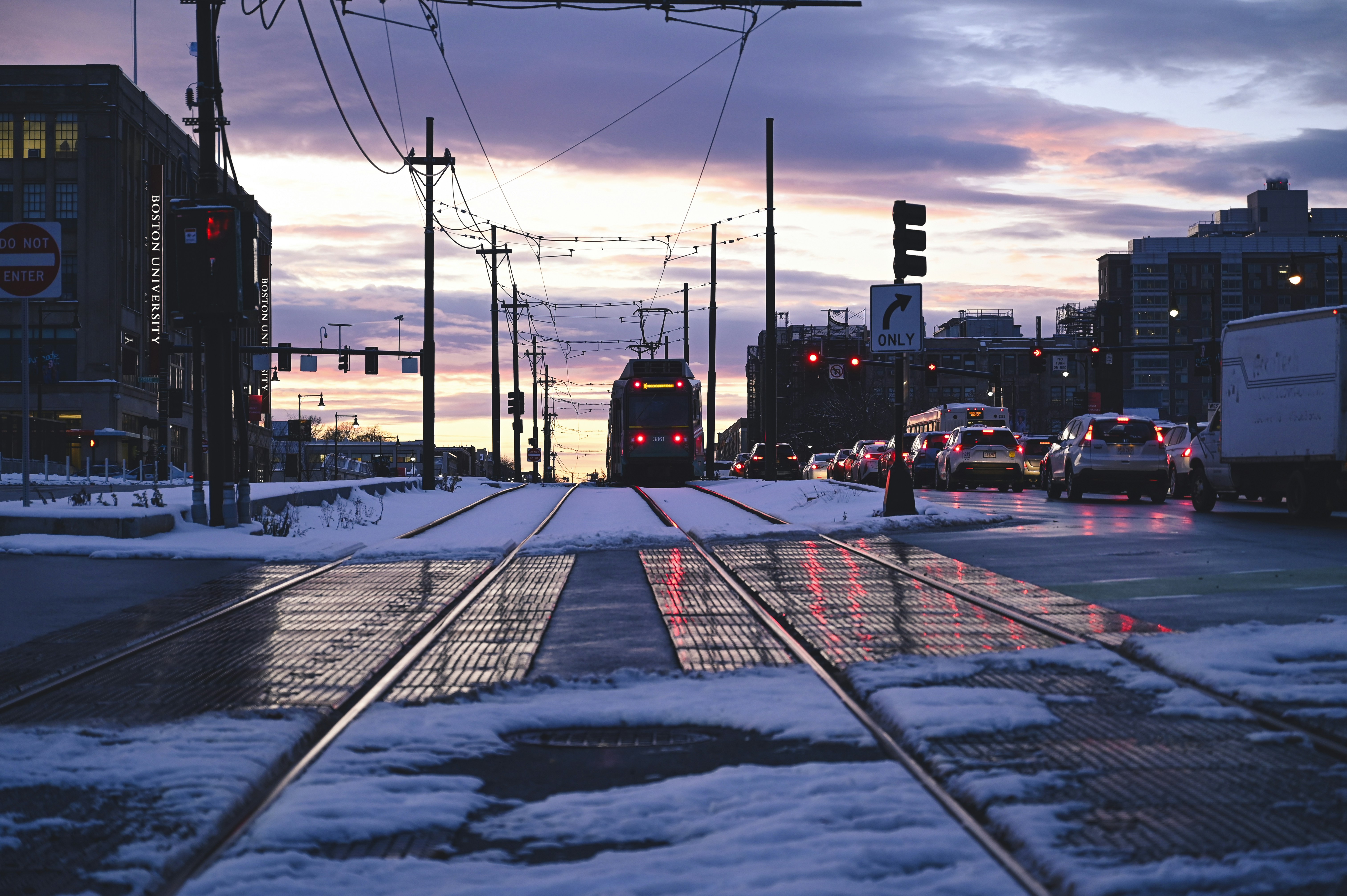 Streetcar approaches under a cloudy, colorful sky with traffic lining a snow-dusted urban street at dusk.