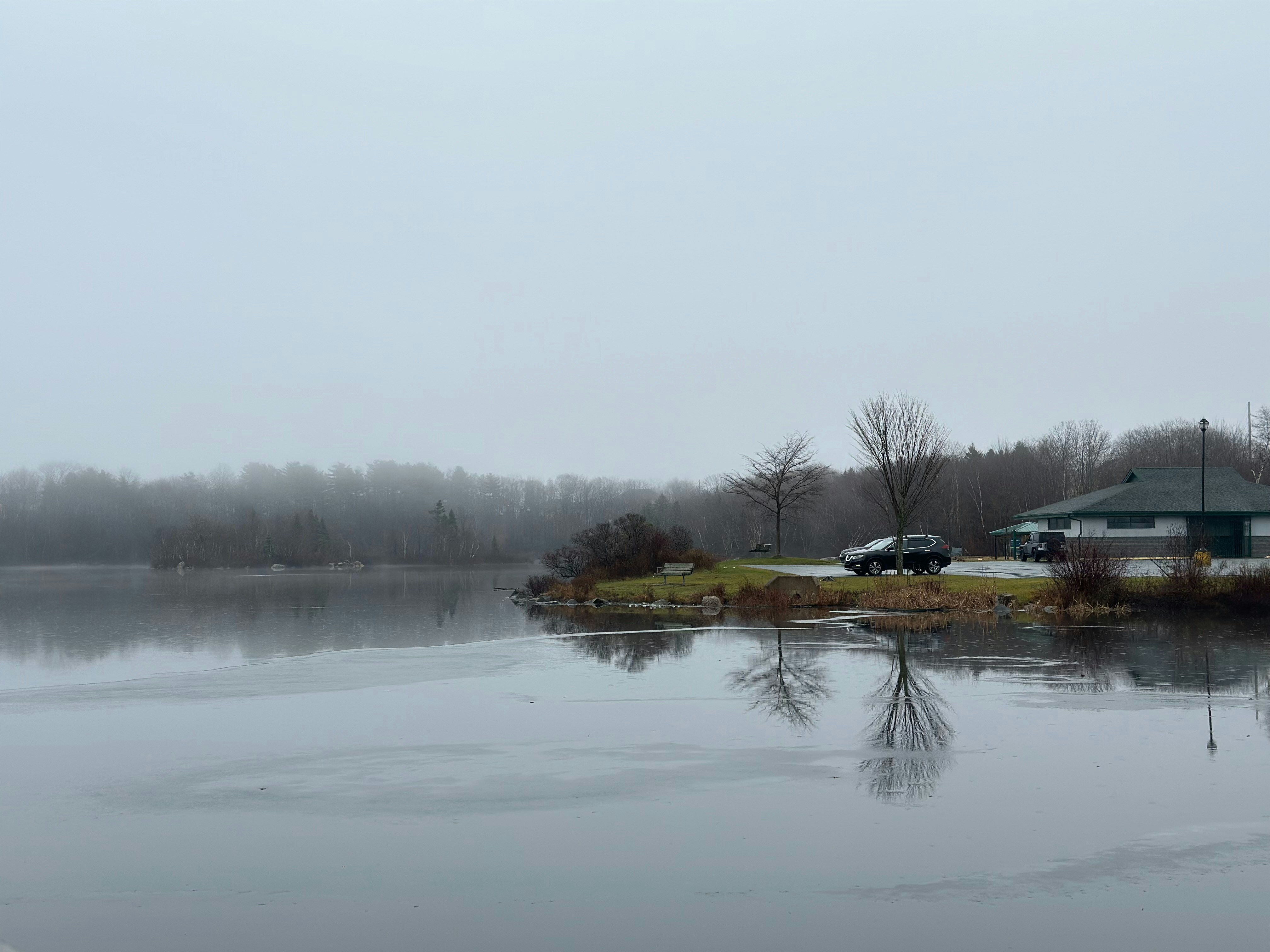 Foggy lake scene with distant trees and a small cabin reflecting in still waters.