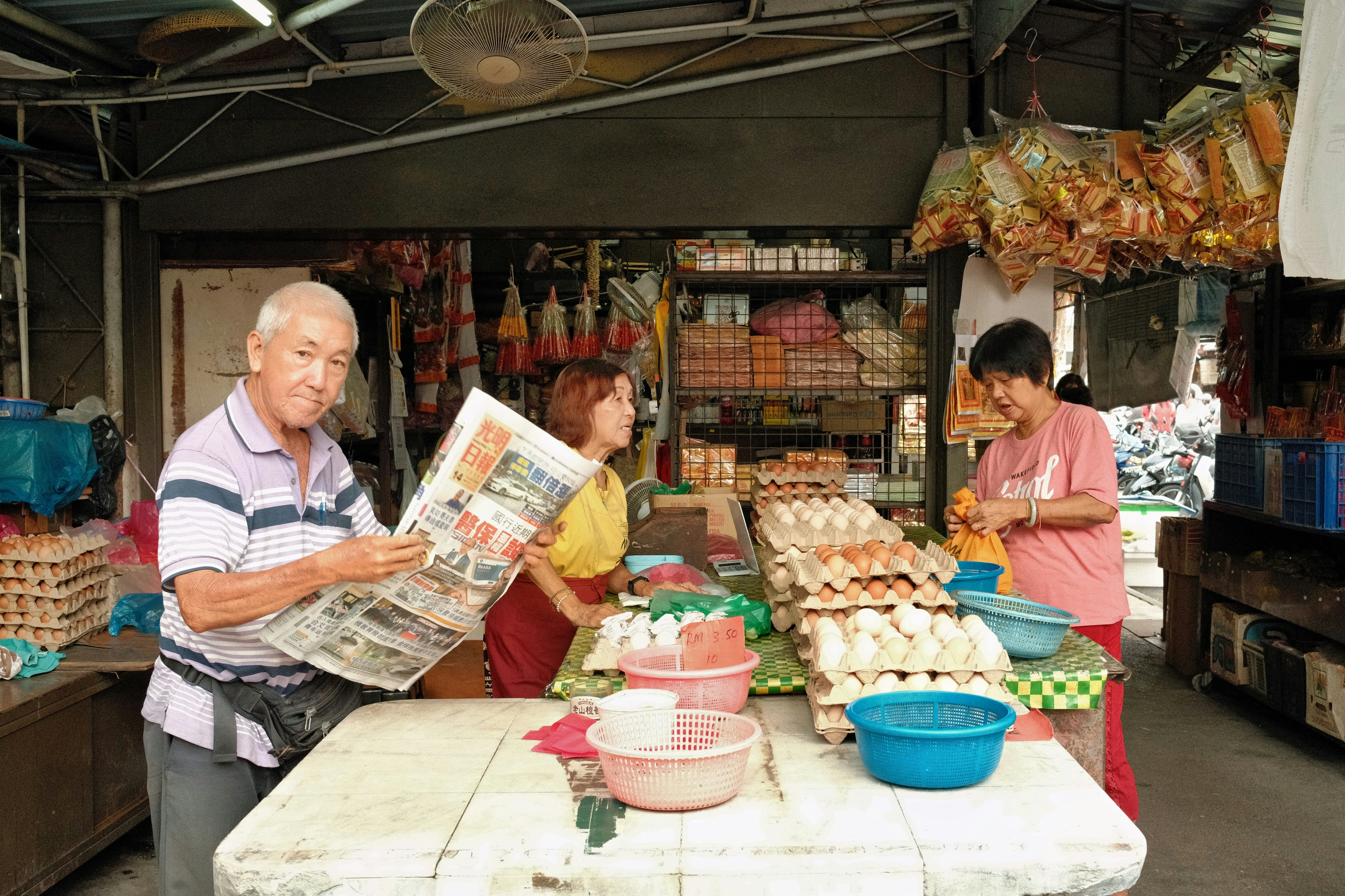 Newspaper Reader at a Lively Market Stall: Quiet Moment Amid Egg Racks