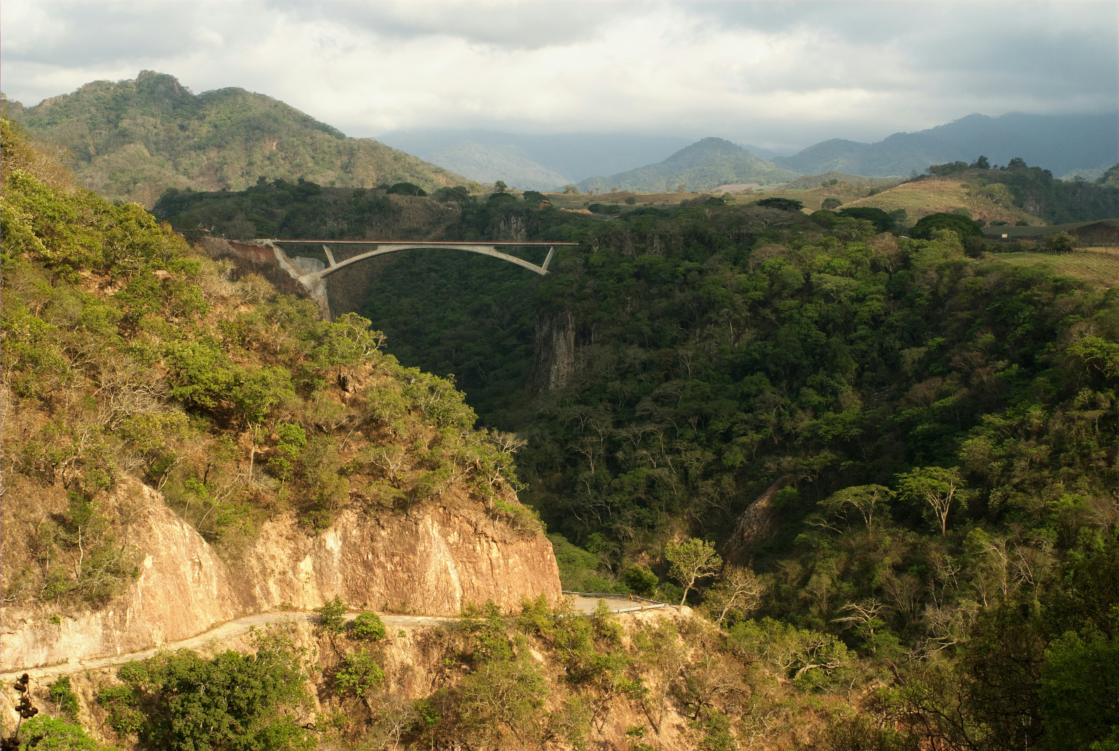 Arching bridge spans a lush, green valley surrounded by rugged hills under a cloudy sky.