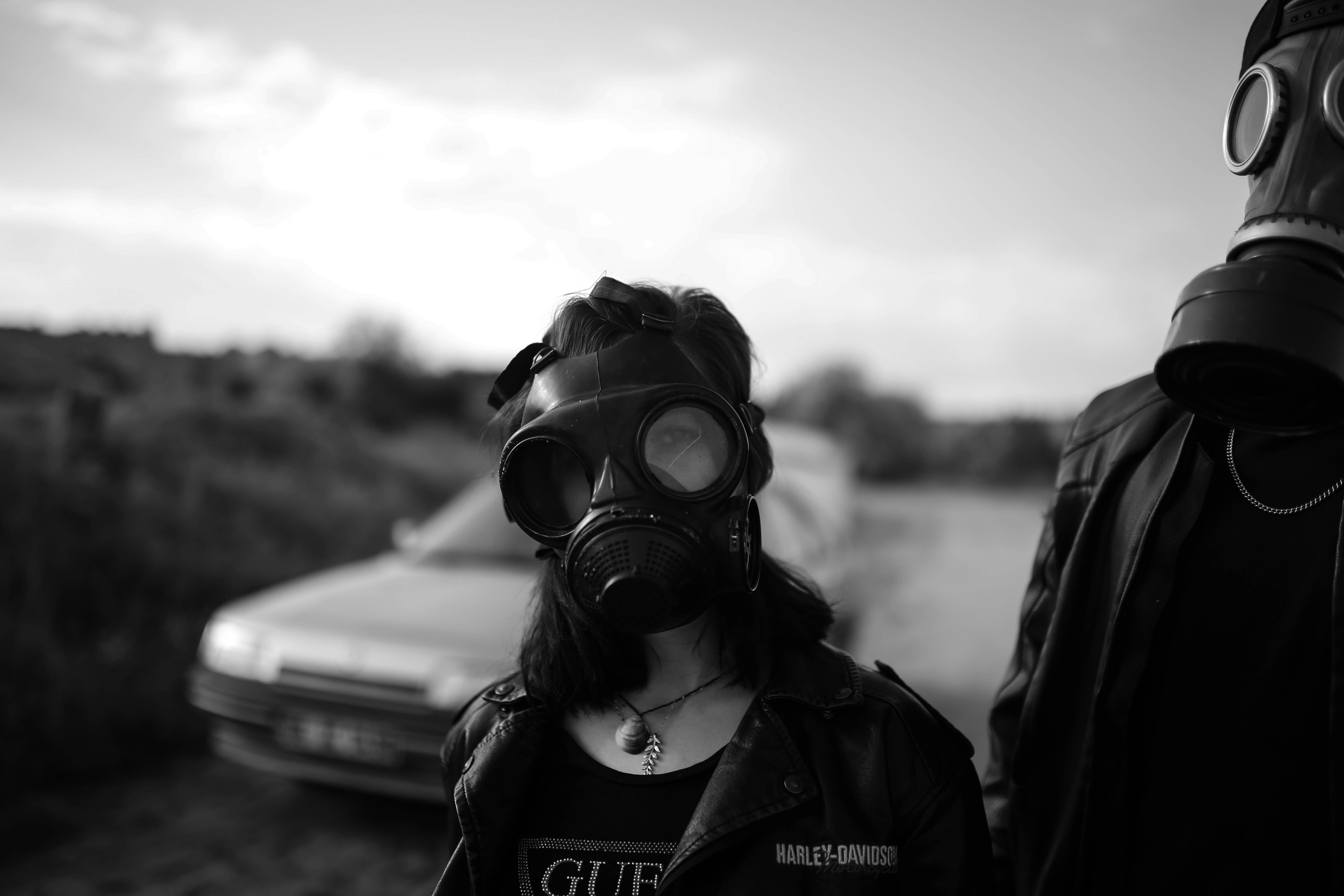 Two individuals wearing gas masks stand amidst an abandoned landscape, hinting at a post-apocalyptic scenario. A vintage car looms in the background.