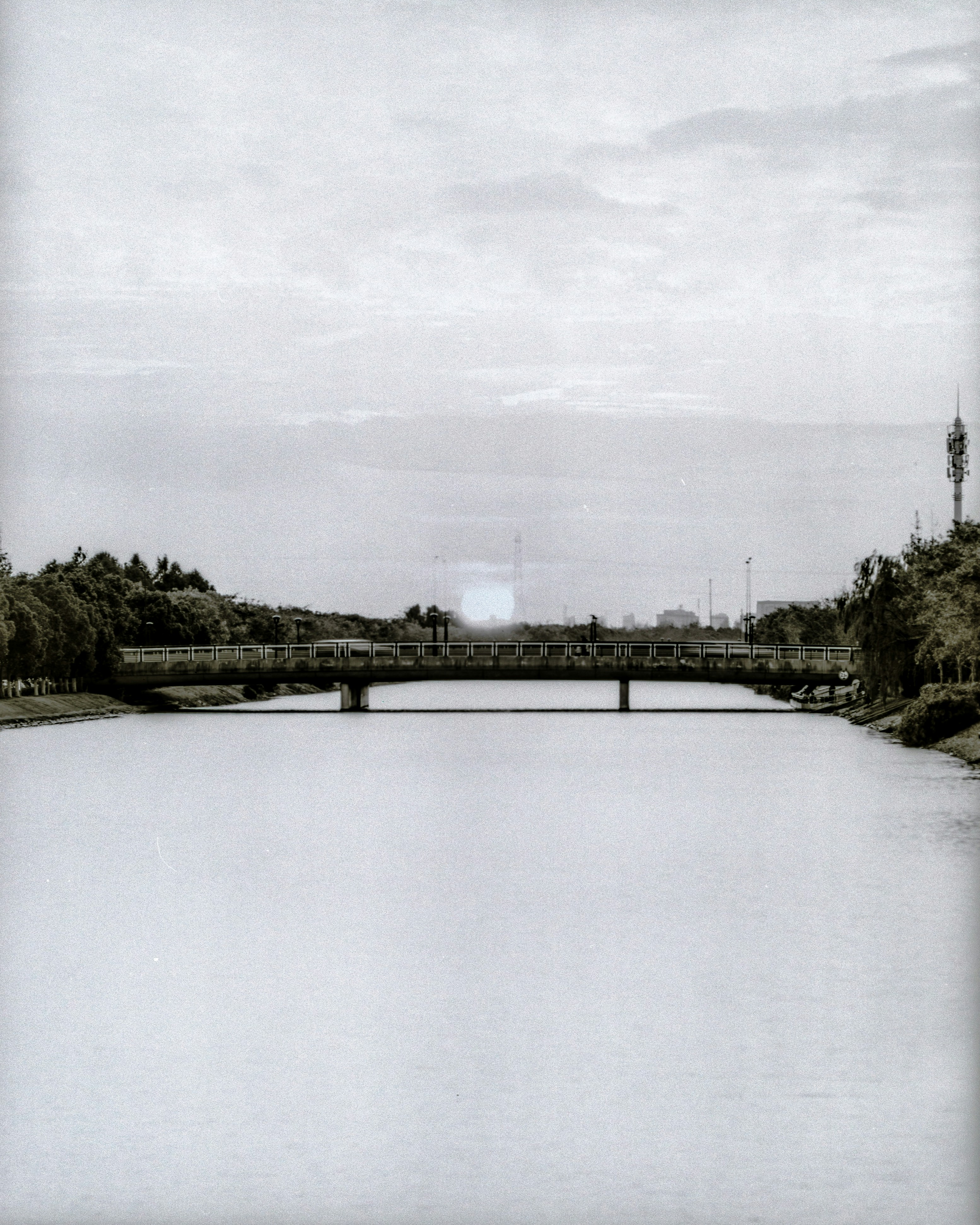 Monochrome photograph of a calm river with a distant bridge spanning the water, framed by trees along the banks.