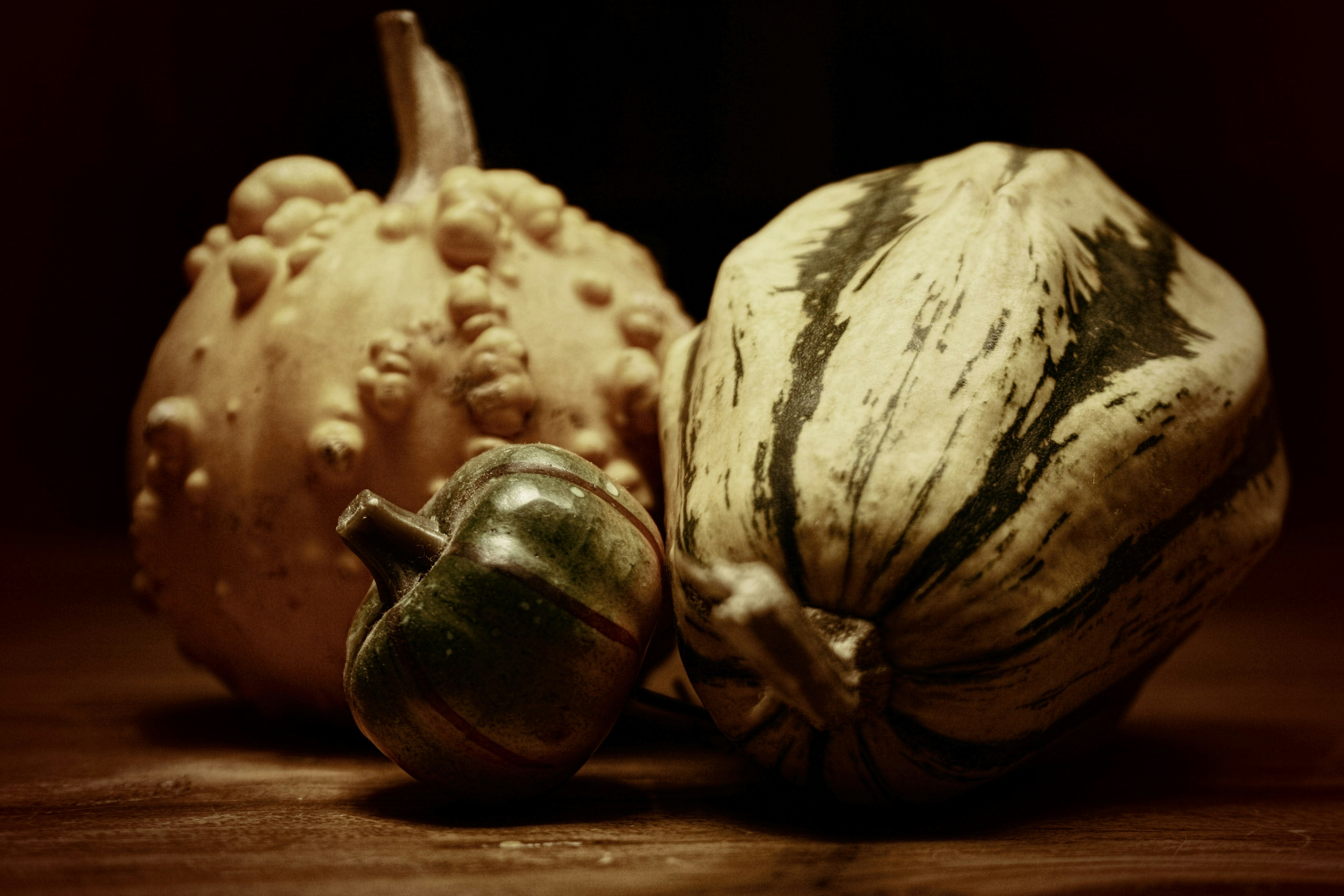 Muted still life of pumpkins and gourds resting on a wooden surface with warm chiaroscuro lighting. The arrangement emphasizes texture and form, creating a contemplative autumn vignette.
