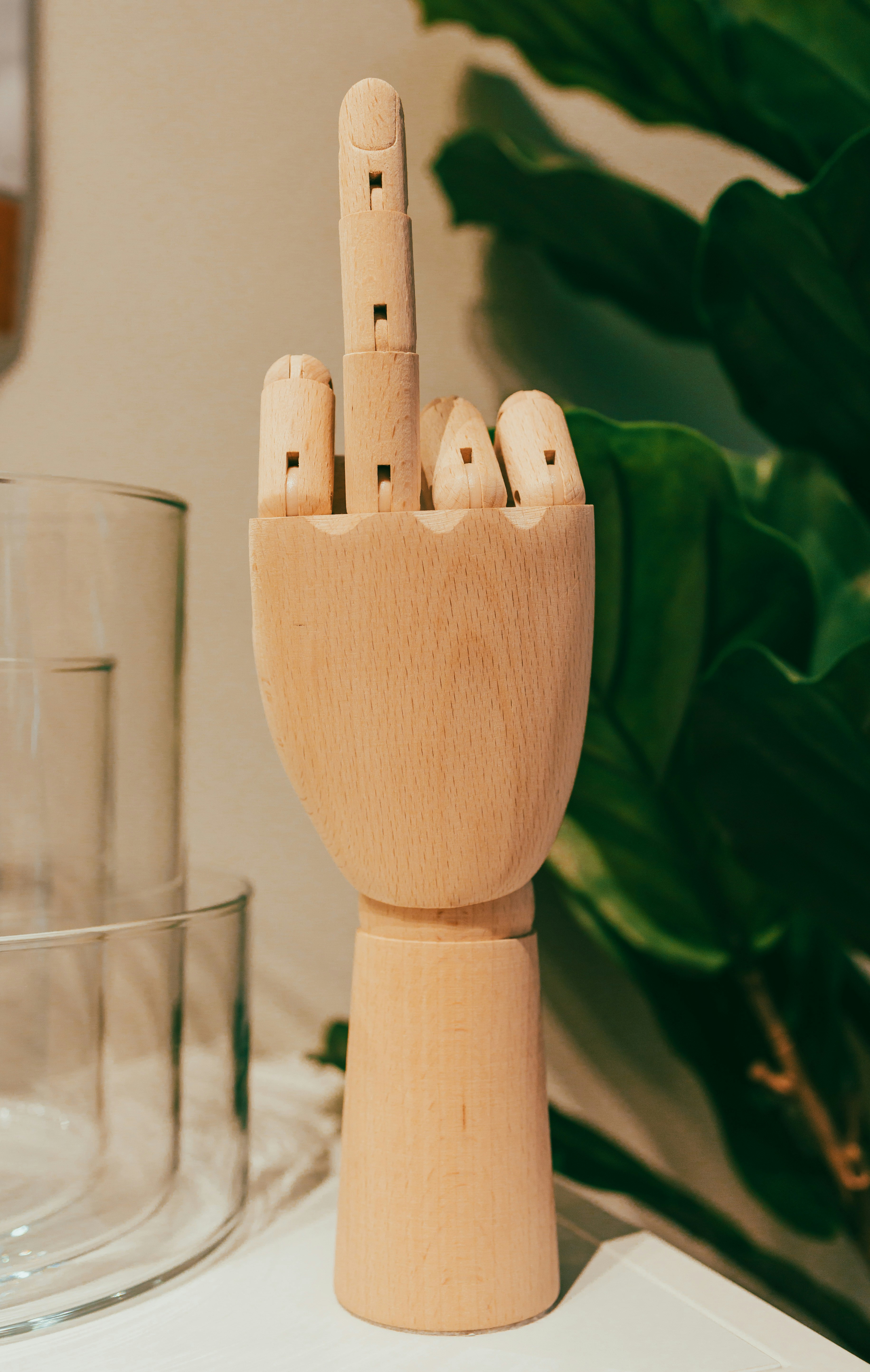 Mid-Finger Still Life: A Wooden Hand Reaches Skyward Among Glass and Greenery
