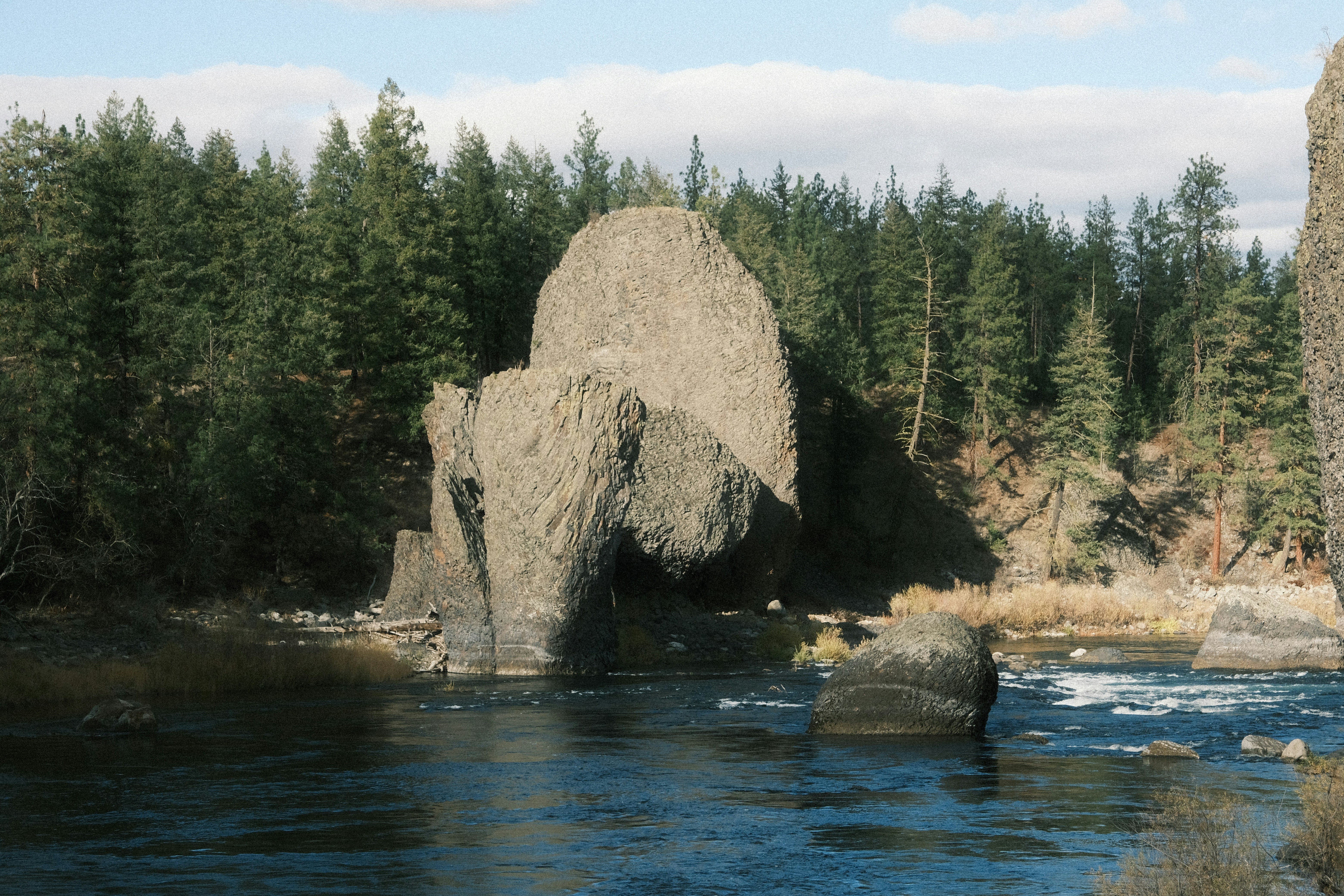 Large rock formations rise from a tranquil river, surrounded by a dense forest under a clear blue sky.
