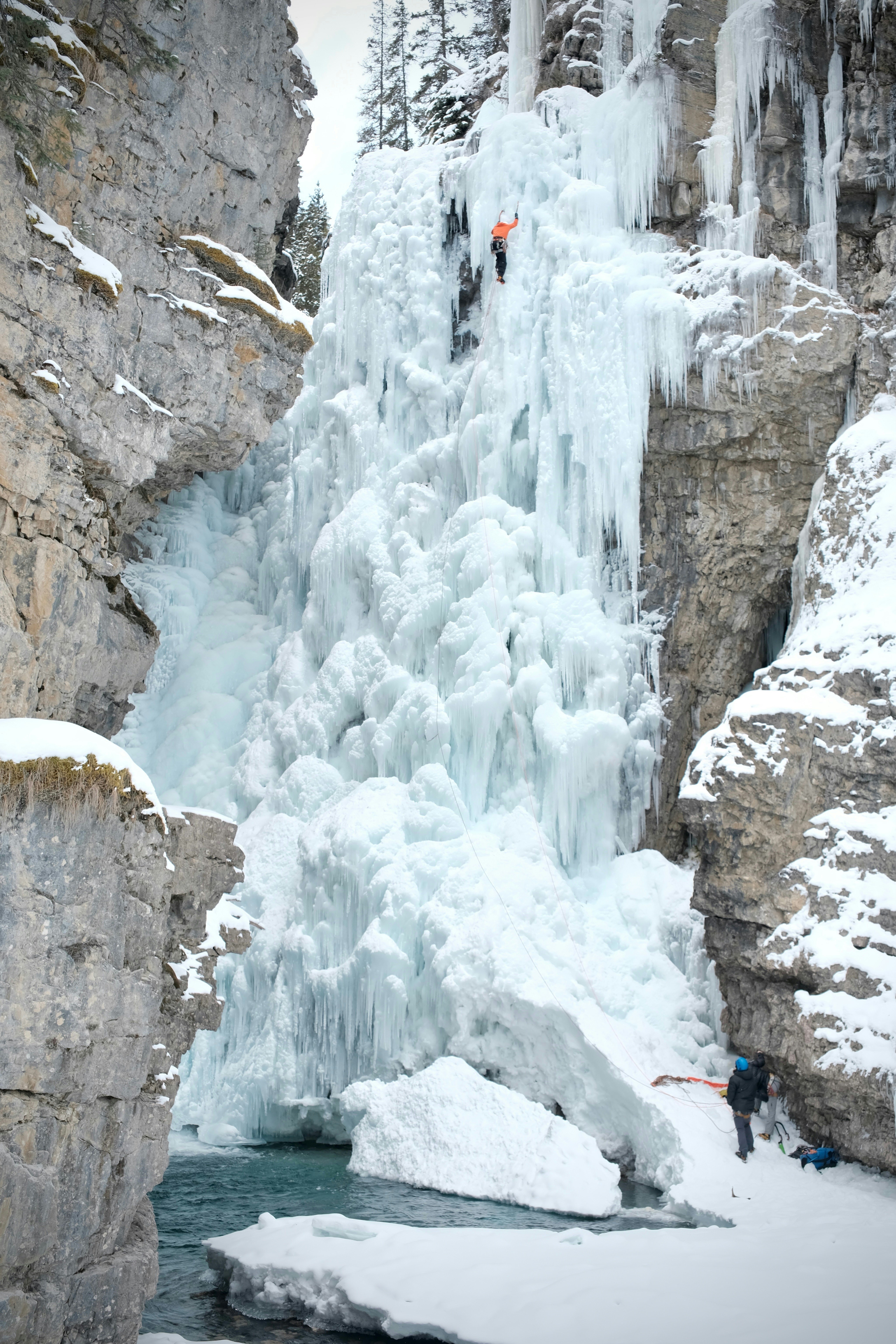 Climber in an orange jacket ascends a towering frozen waterfall amid rugged cliffs, with icy blues contrasting against earthy rock tones.