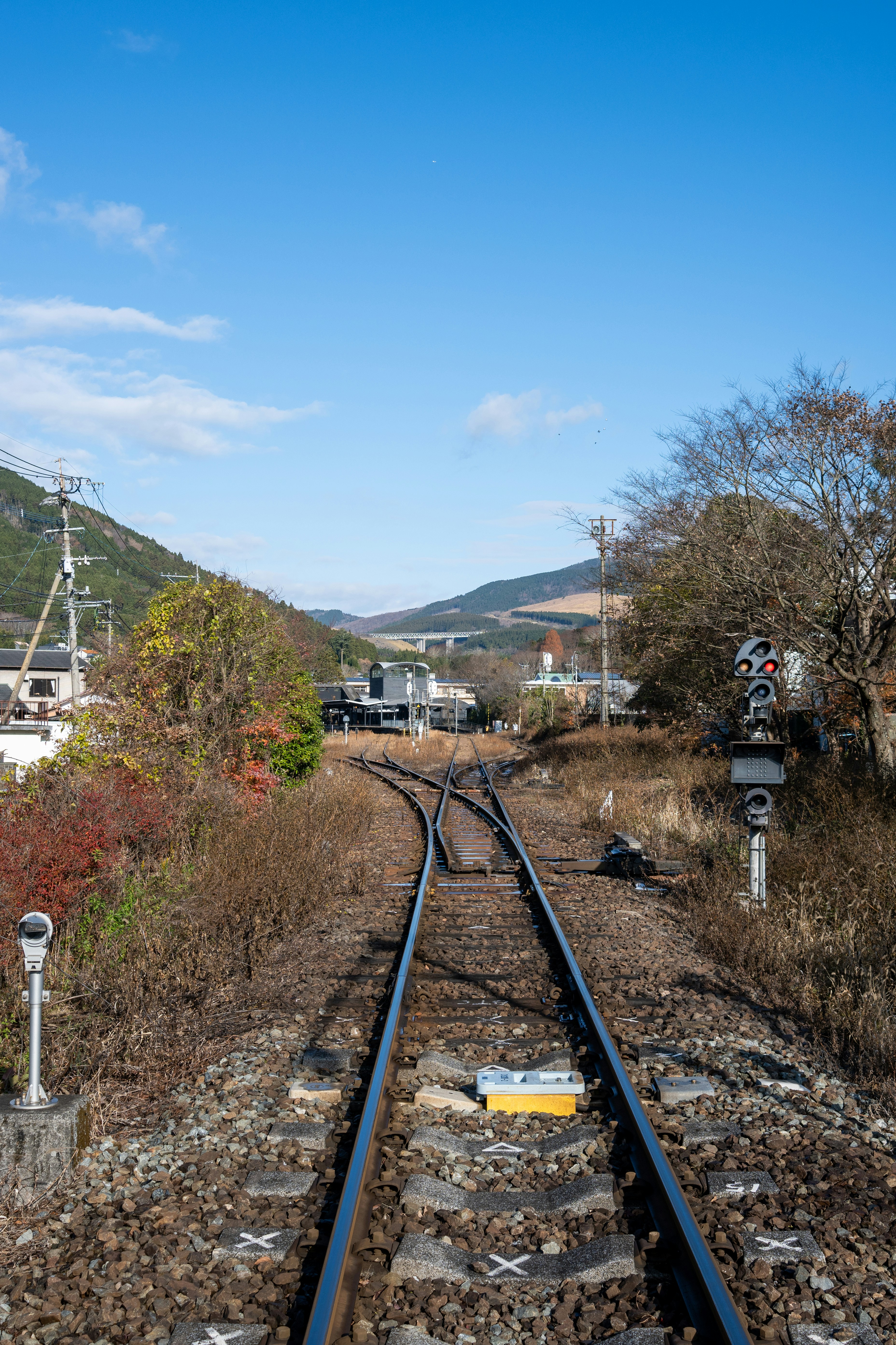 Railway tracks curve toward distant hills, flanked by scrub and a signaling post on the right. The scene captures a quiet, rural rail corridor under a clear blue sky.