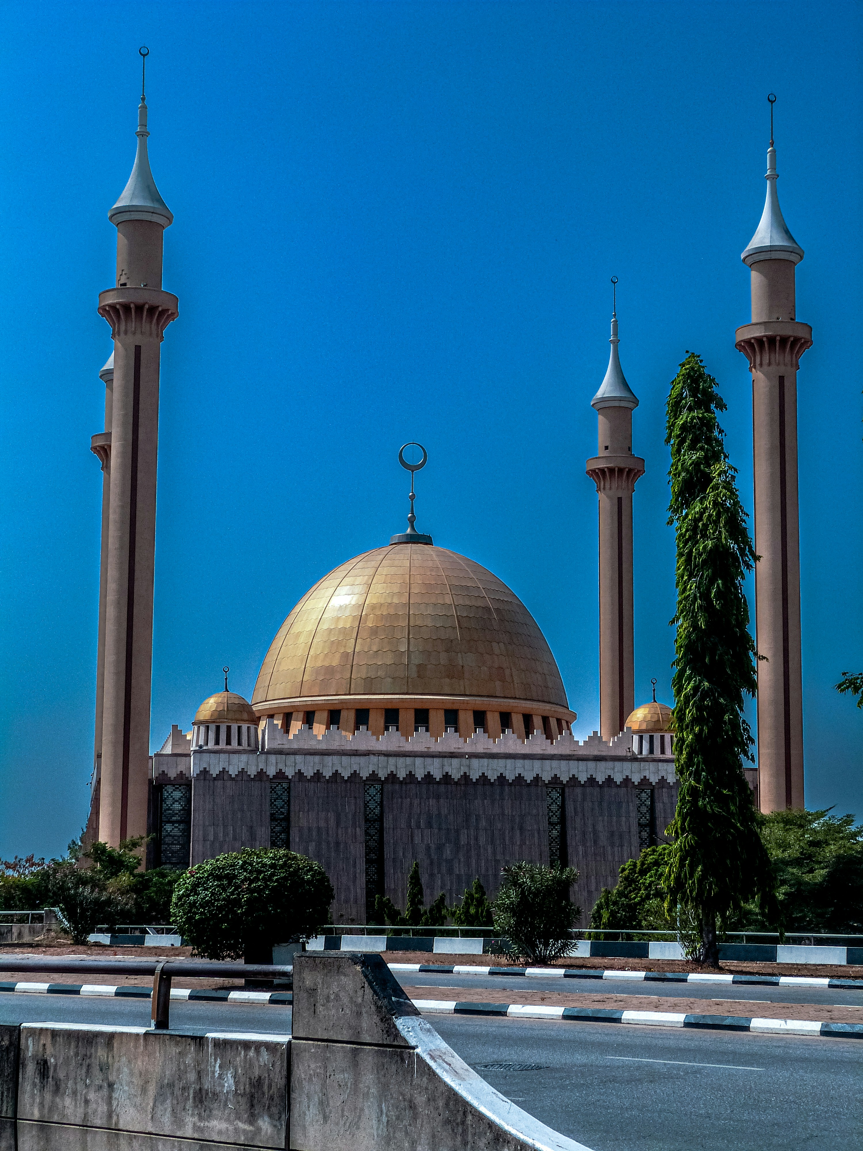 A mosque with a gleaming gold dome and slender minarets stands against a vivid blue sky, with landscaped foreground and a paved approach.