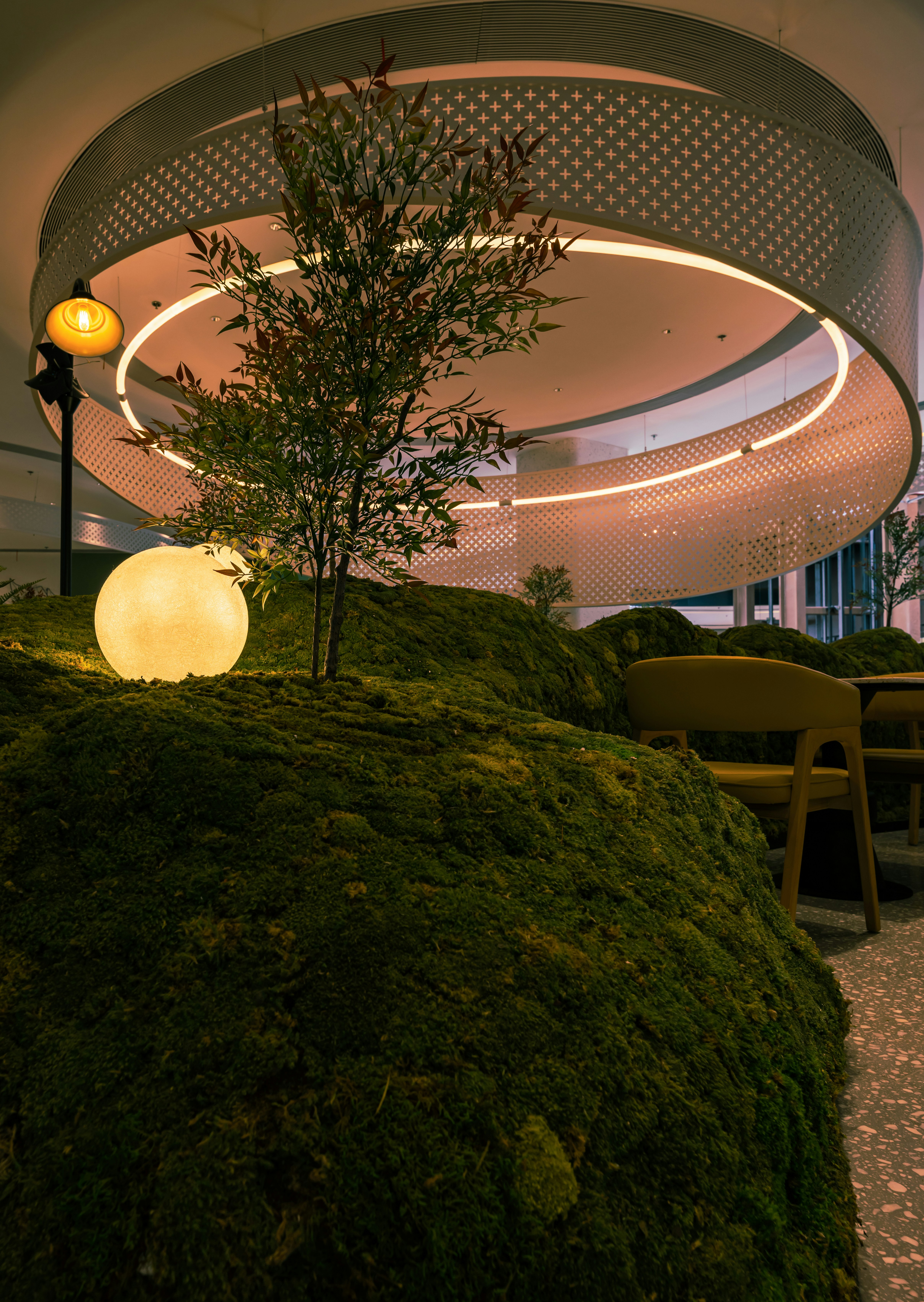 Moss-covered hill with a glowing apple-shaped lamp sits under a circular perforated ceiling and neon ring, inside a contemporary lounge.