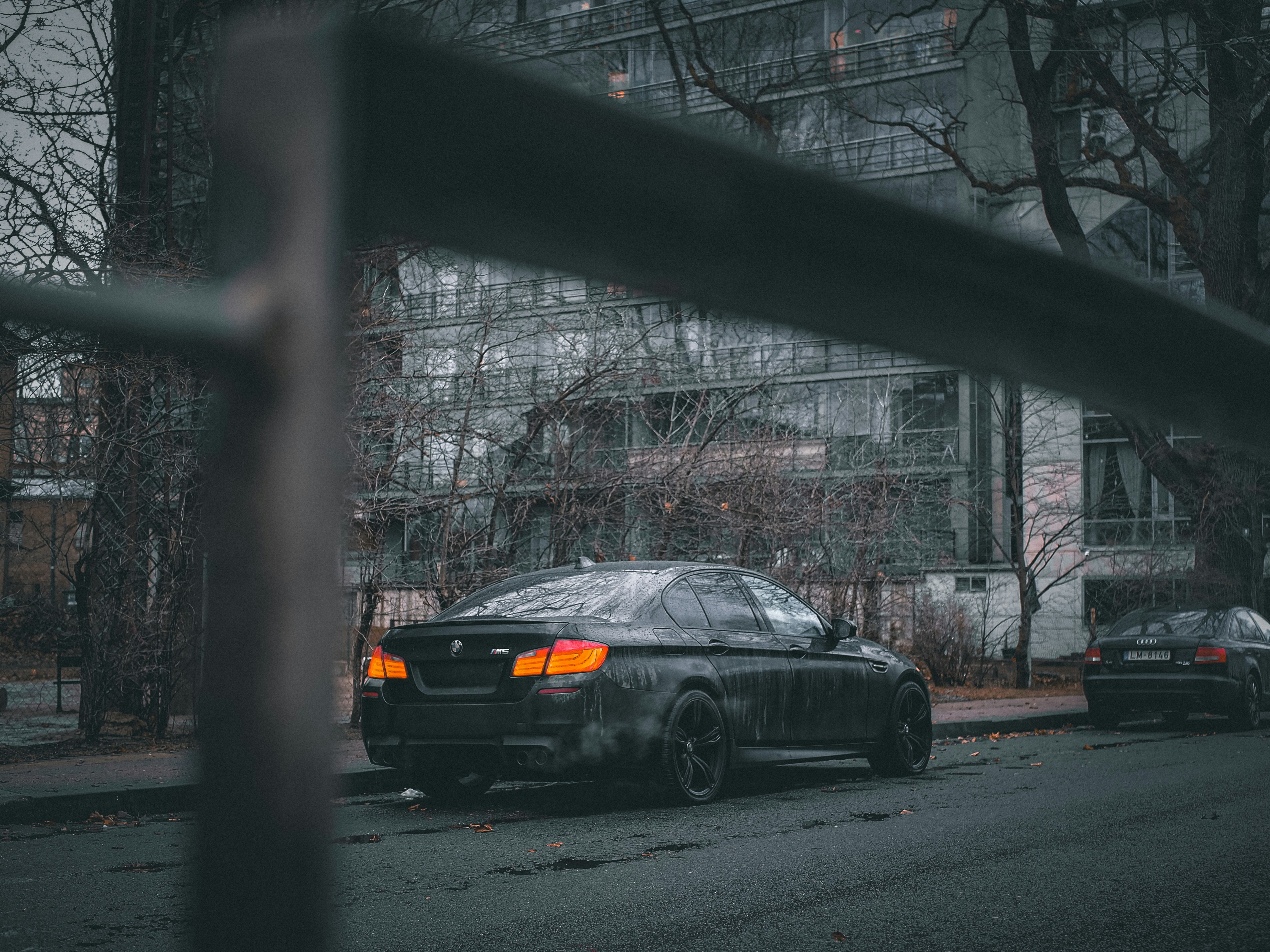 Sleek black car parked on a city street, partially obscured by a blurred metal railing, with a modern building in the background.