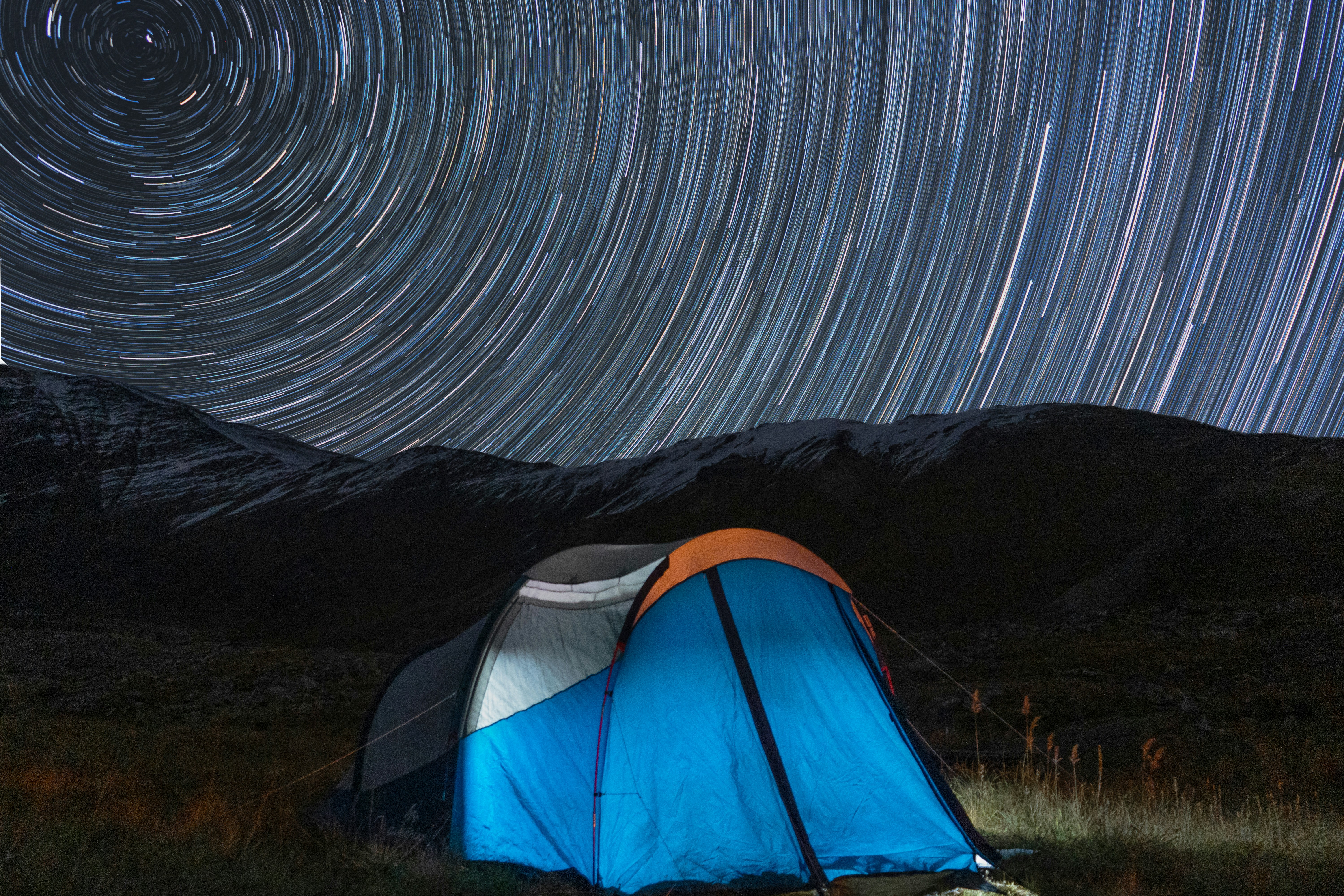 Vibrant blue tent glows beneath swirling star trails in a night sky, framed by the silhouette of distant mountains.