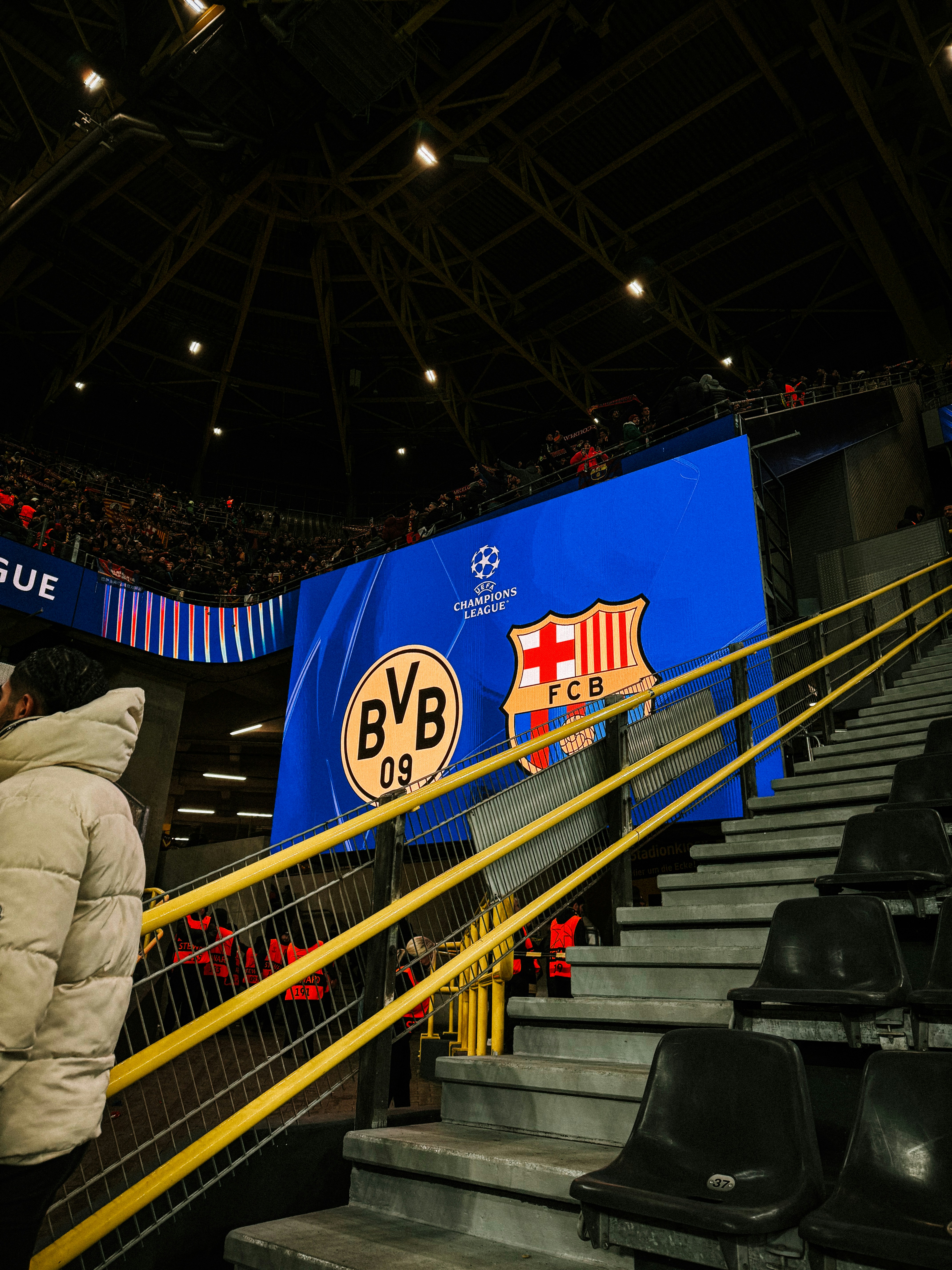 Football fans gather in a stadium with a display showing Borussia Dortmund and FC Barcelona logos.