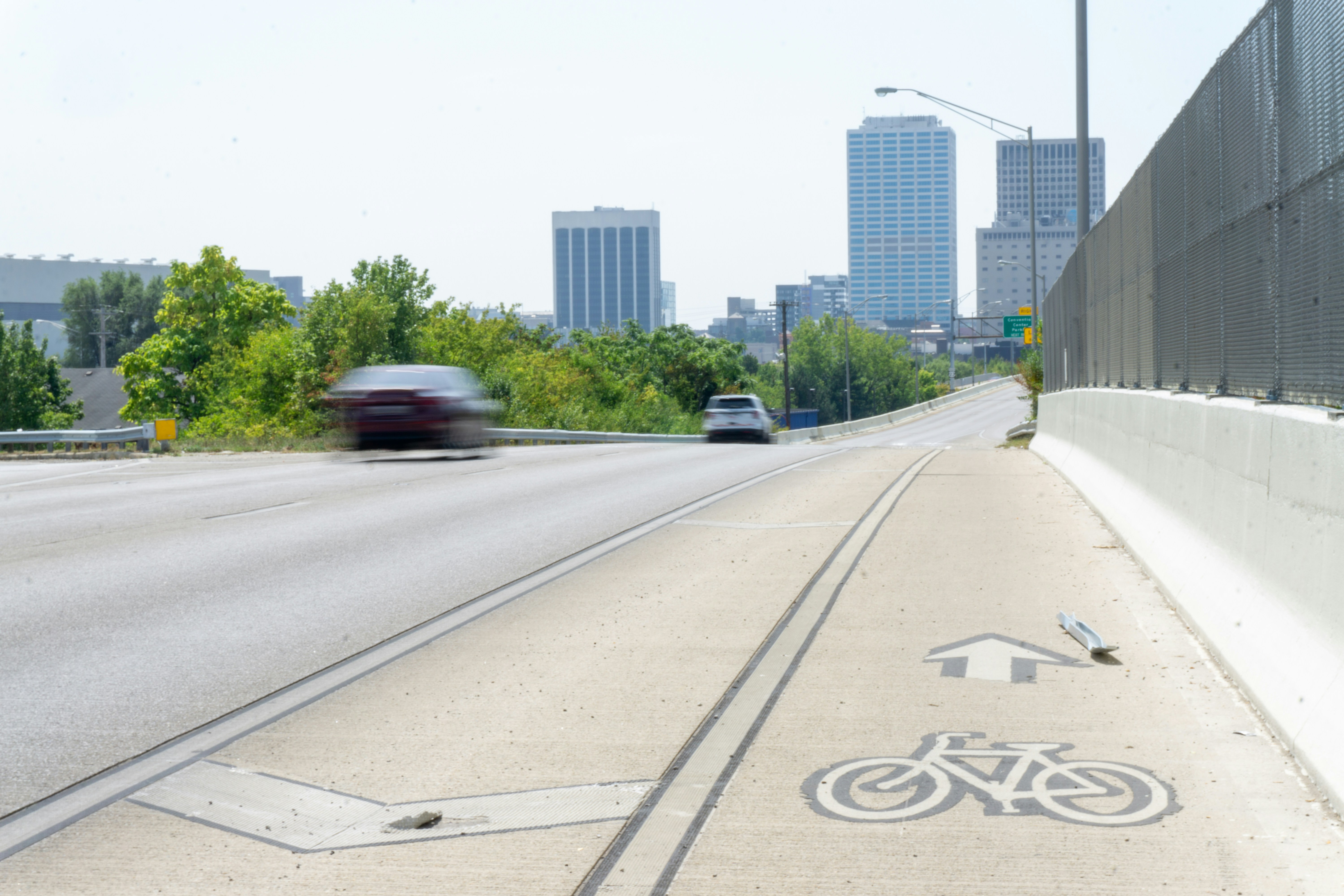 Bike lane runs alongside a highway with blurred cars and distant city skyline under a clear sky.