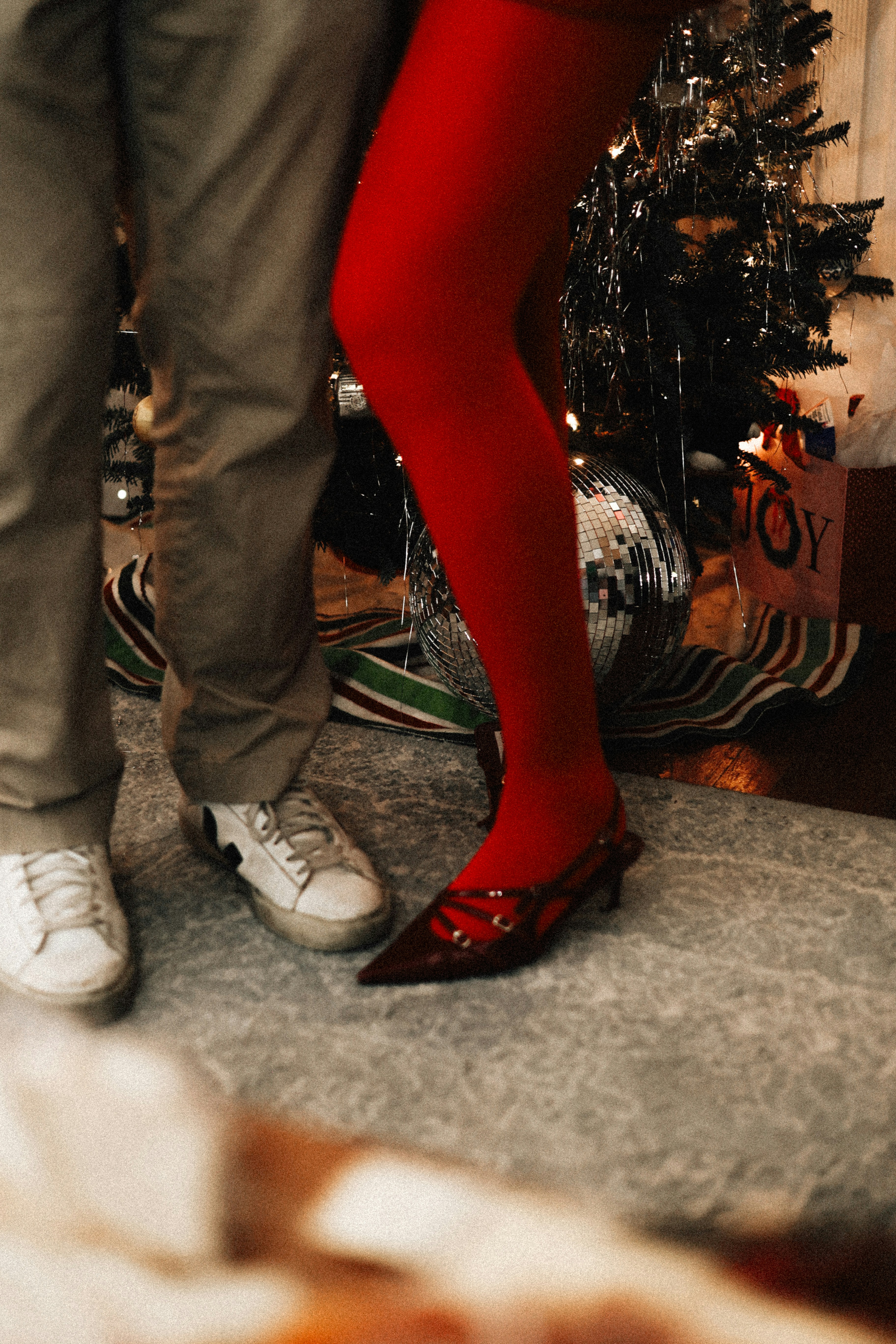 A close-up of a person's red legwear paired with stylish shoes, set against a backdrop of holiday decorations and a disco ball. The scene exudes a cheerful, festive atmosphere.