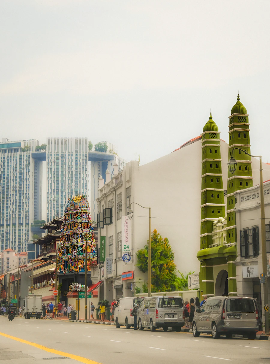 Vibrant sculpted Hindu gopuram rising along a city street, framed by green mosque towers and modern high-rise buildings.