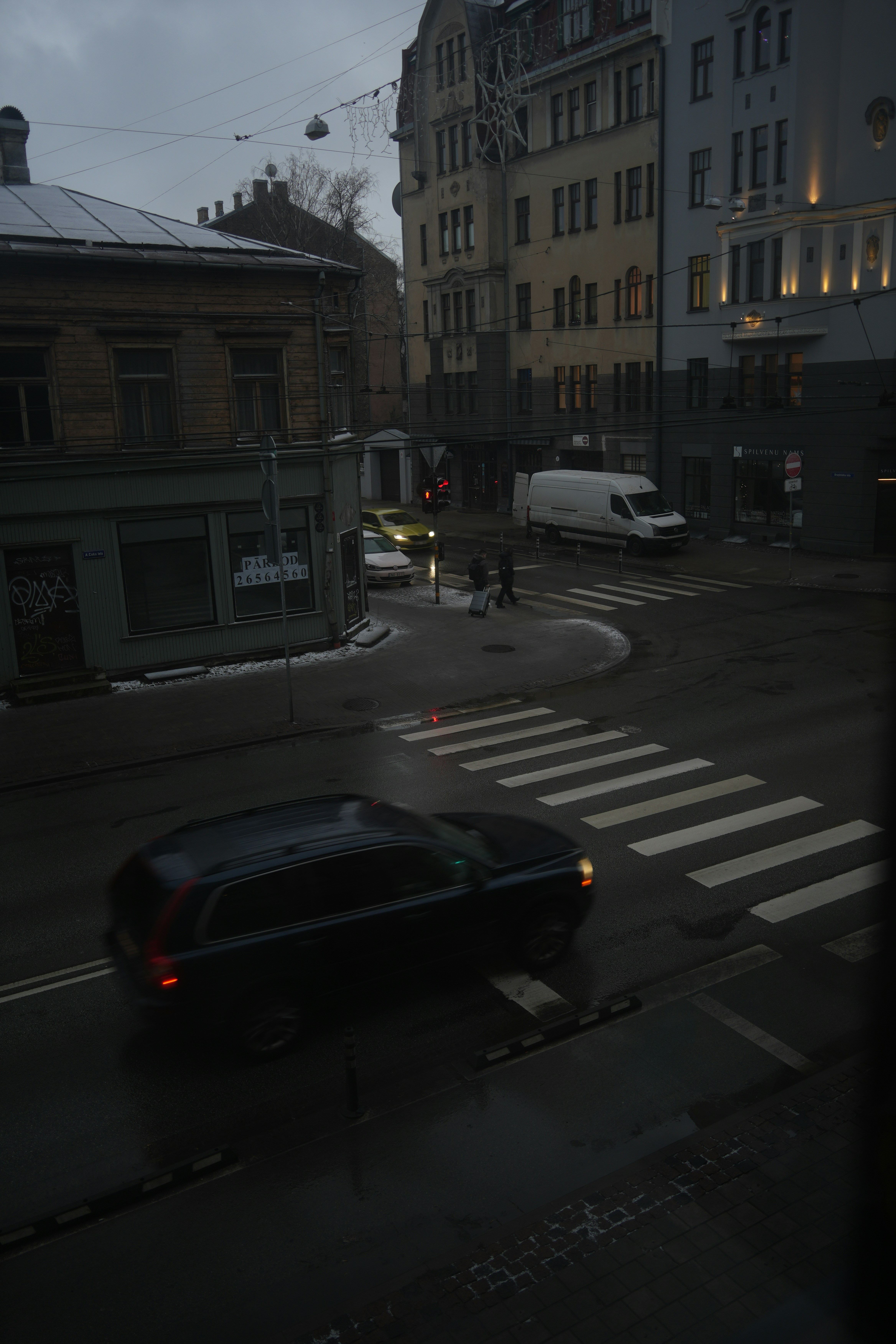 A gloomy urban scene featuring a rainy city street corner. A black SUV crosses a wet pedestrian crosswalk while pedestrians walk by with umbrellas and luggage. The dim light and reflections on the wet pavement highlight the cold, moody atmosphere of the city, surrounded by old buildings and a quiet sense of movement.