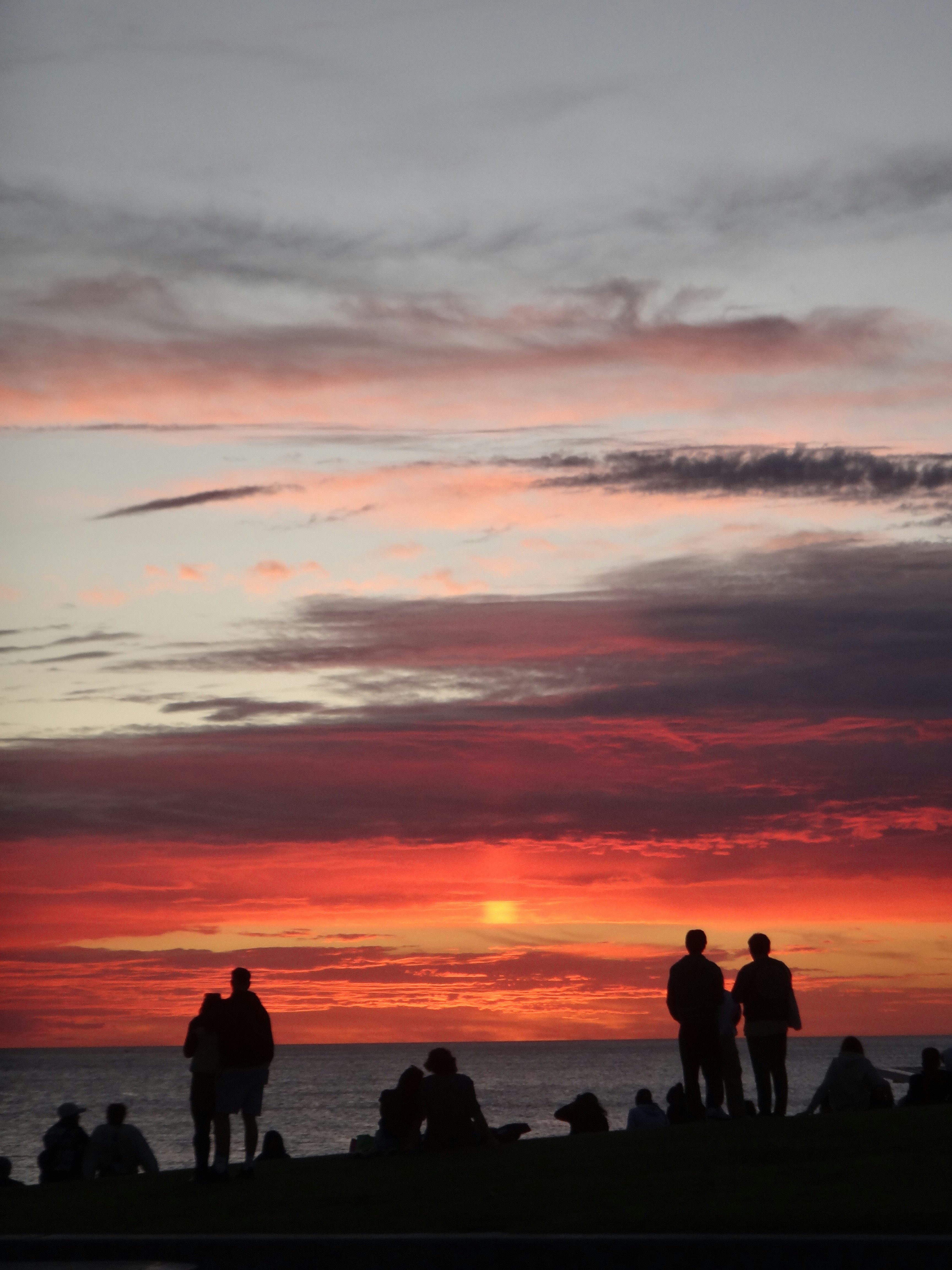 Silhouettes of people on a grassy coastal dune against a fiery sunset over the calm sea.