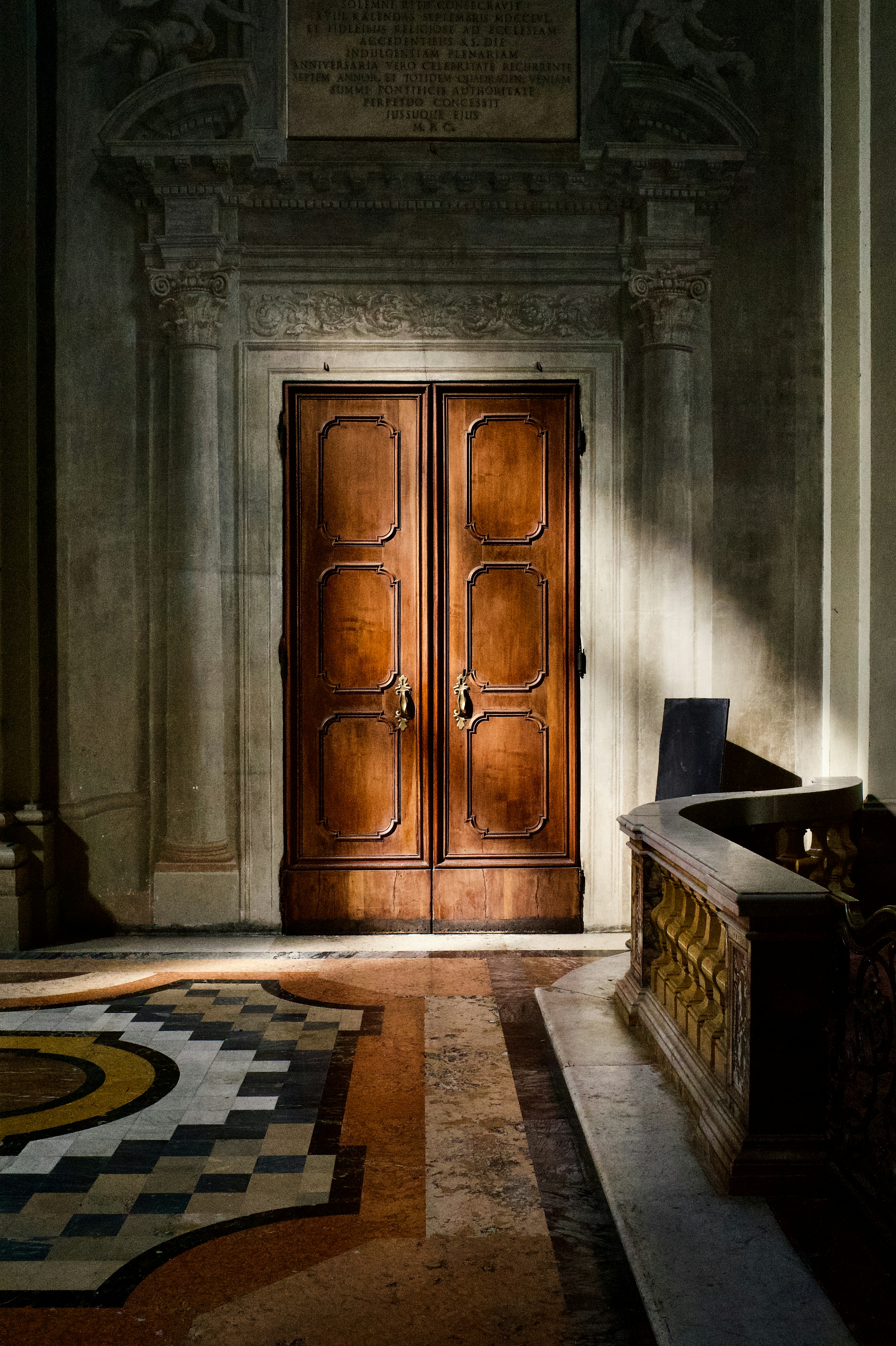 Ornate wooden door illuminated by a soft beam of light, enhancing its intricate carvings and warm brown hues. The surrounding stone architecture and colorful mosaic floor create a harmonious interplay of light and shadow.