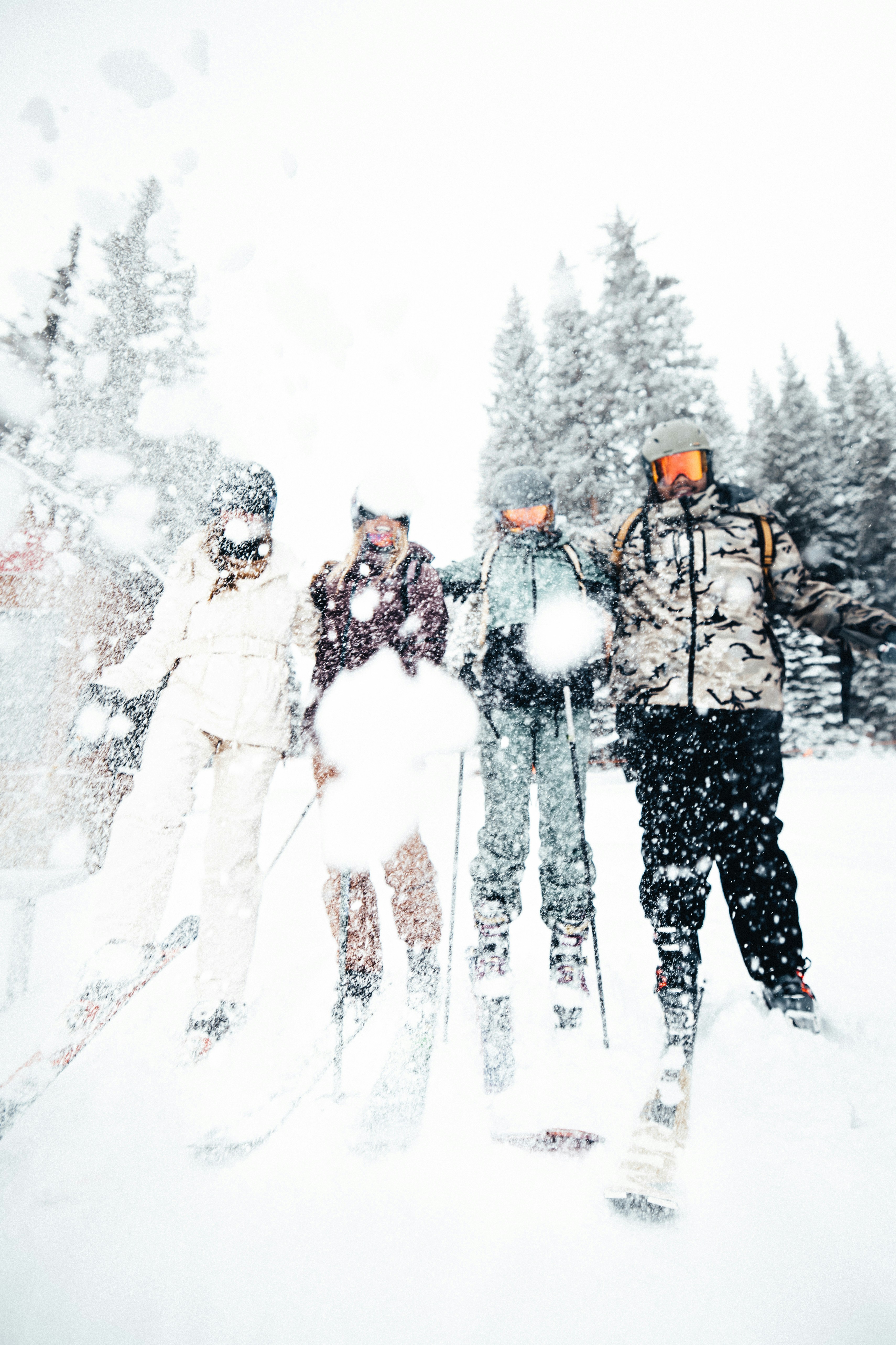Four skiers in winter gear with reflective goggles surrounded by falling snow in a snowy forest.