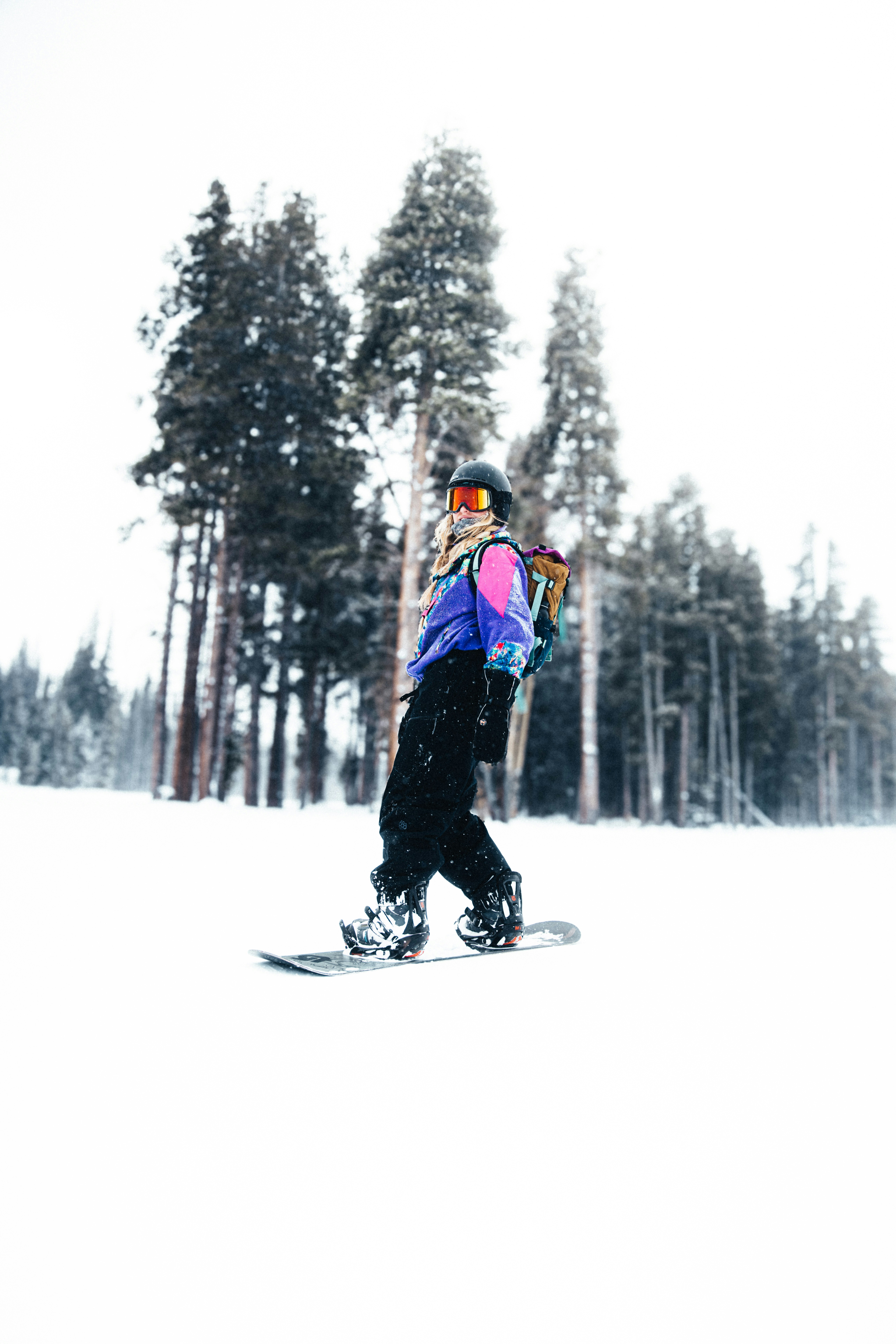 Snowboarder in vibrant purple and pink attire glides through snow, surrounded by tall evergreen trees.
