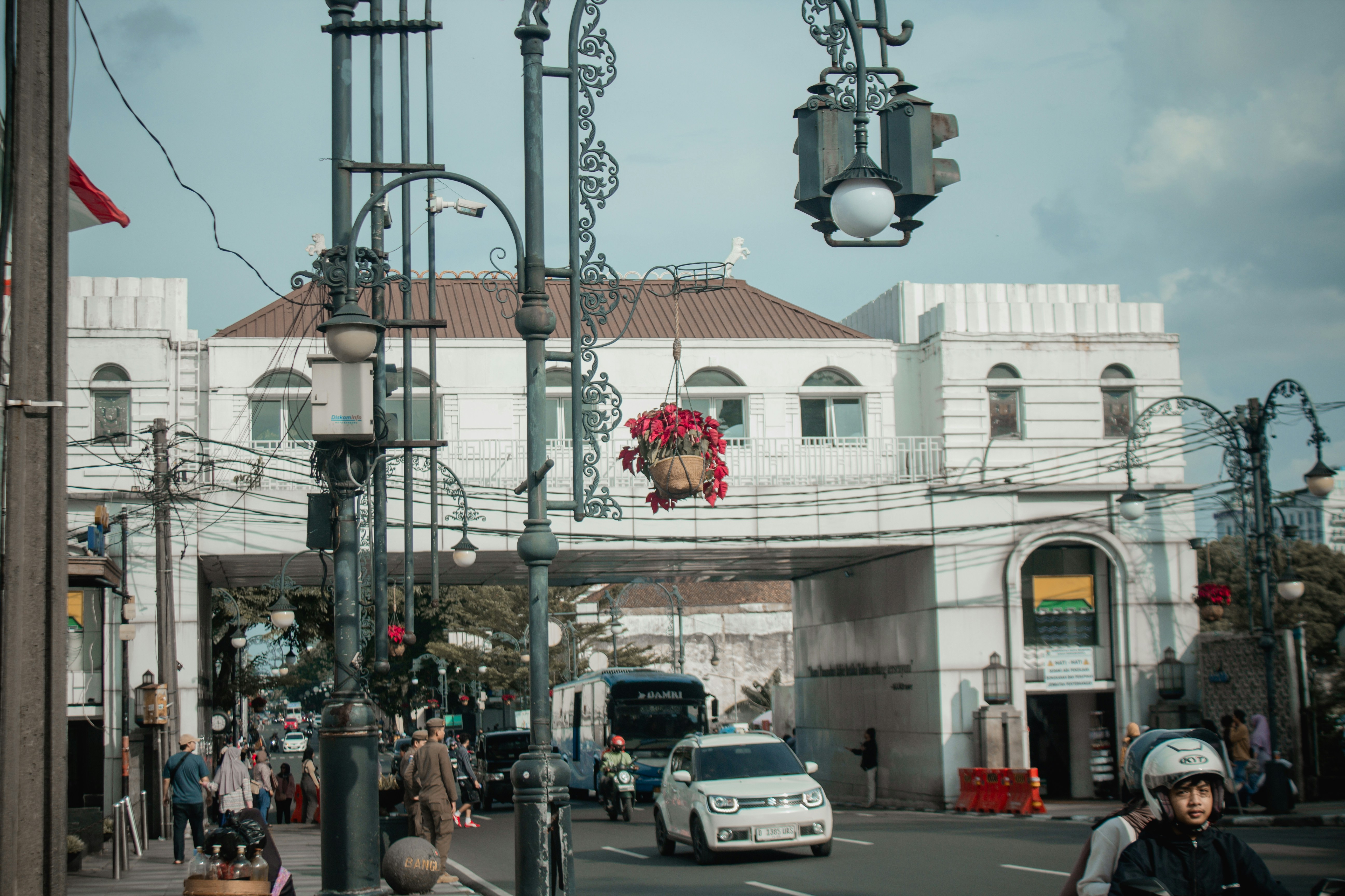 This vibrant street scene in Bandung showcases the charming blend of modernity and tradition, highlighted by decorative streetlamps and colorful flower baskets. The iconic architecture in the background adds a historical touch, while pedestrians and vehicles navigate the bustling thoroughfare, embodying the lively spirit of urban life.