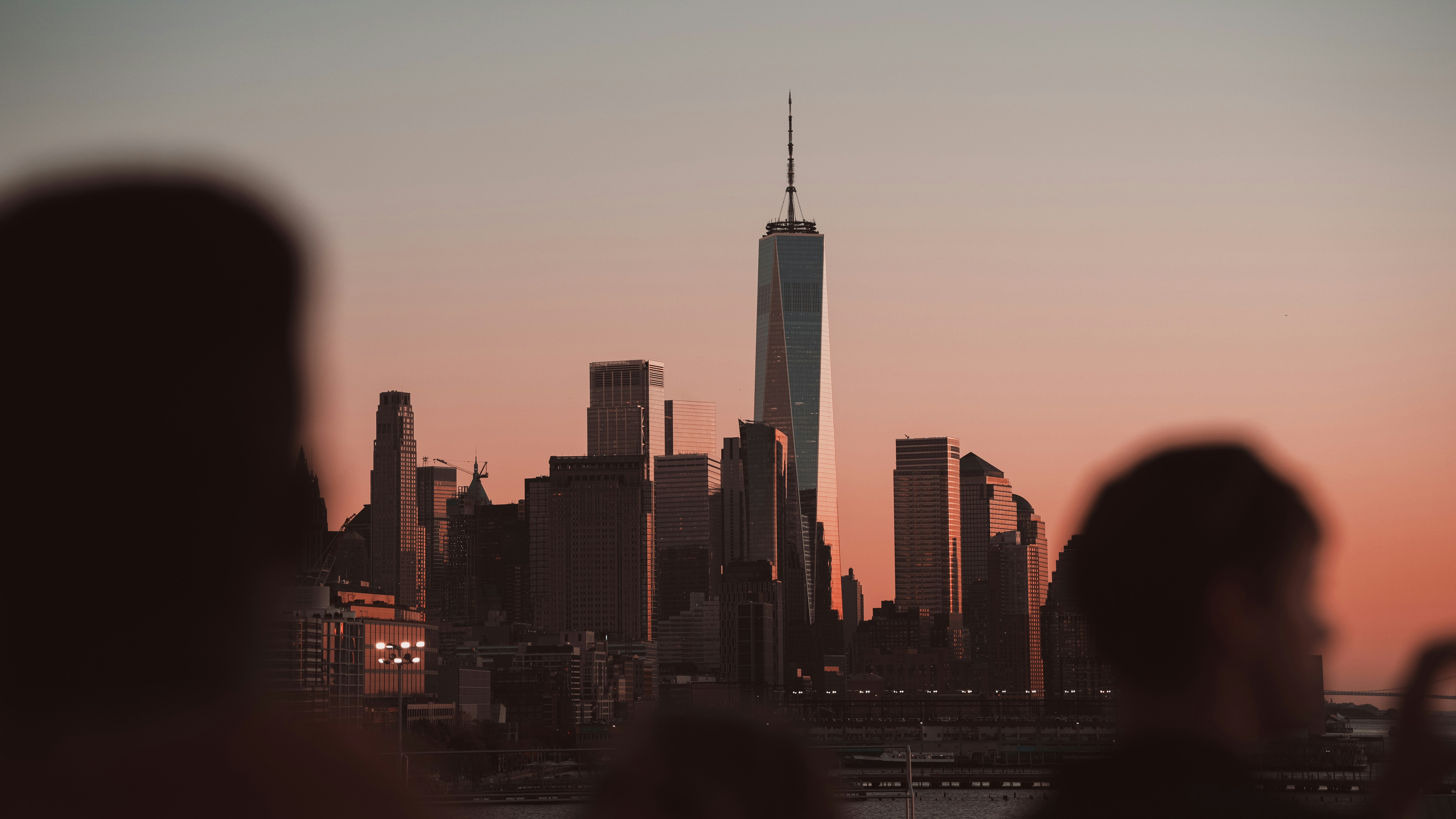City skyline at sunset with One World Trade Center centered against an orange-pink sky. Foreground silhouettes of people add depth to the scene.