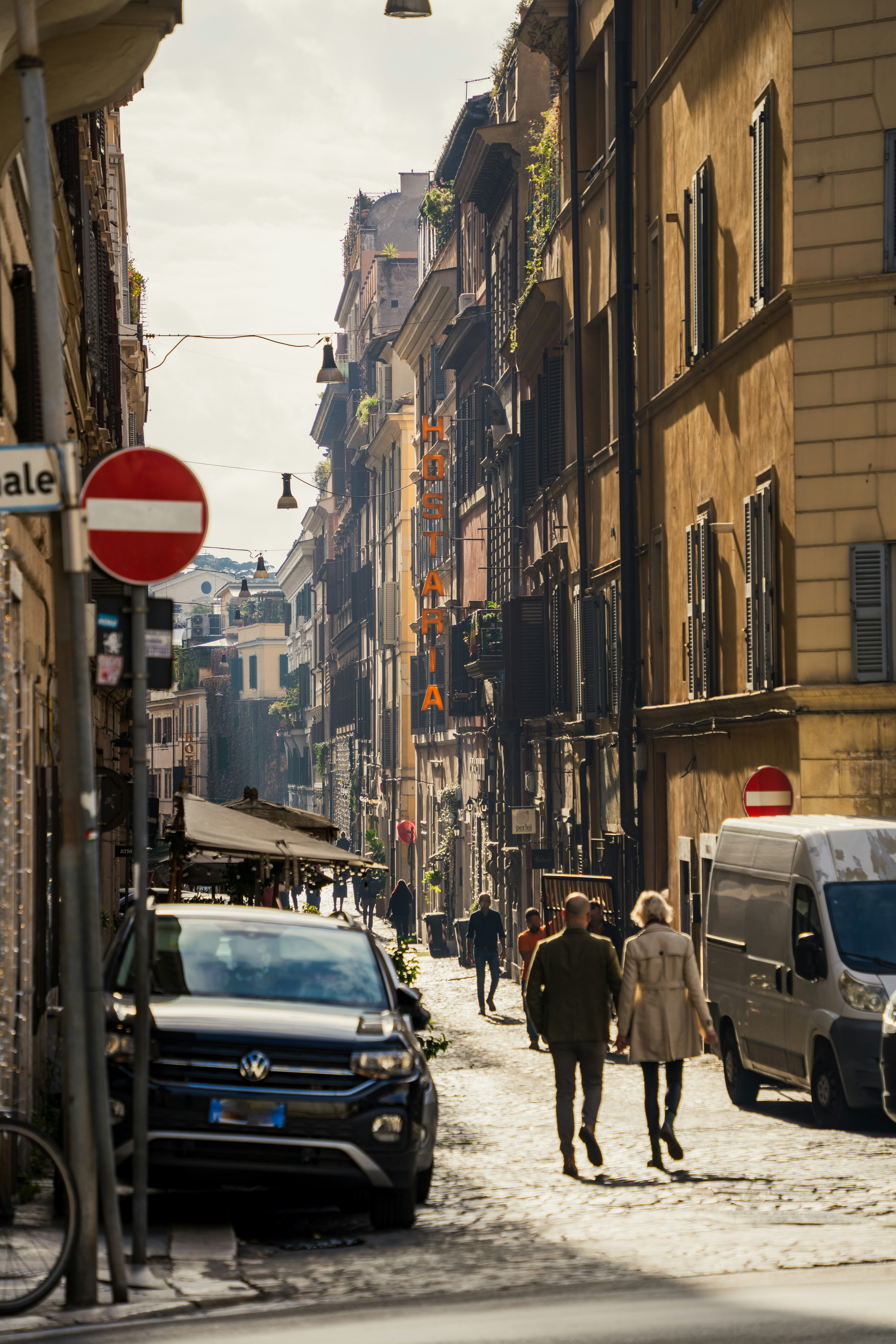 Golden sunlight bathes a cobblestone street lined with historic buildings in Rome, as pedestrians walk past parked cars and vans.